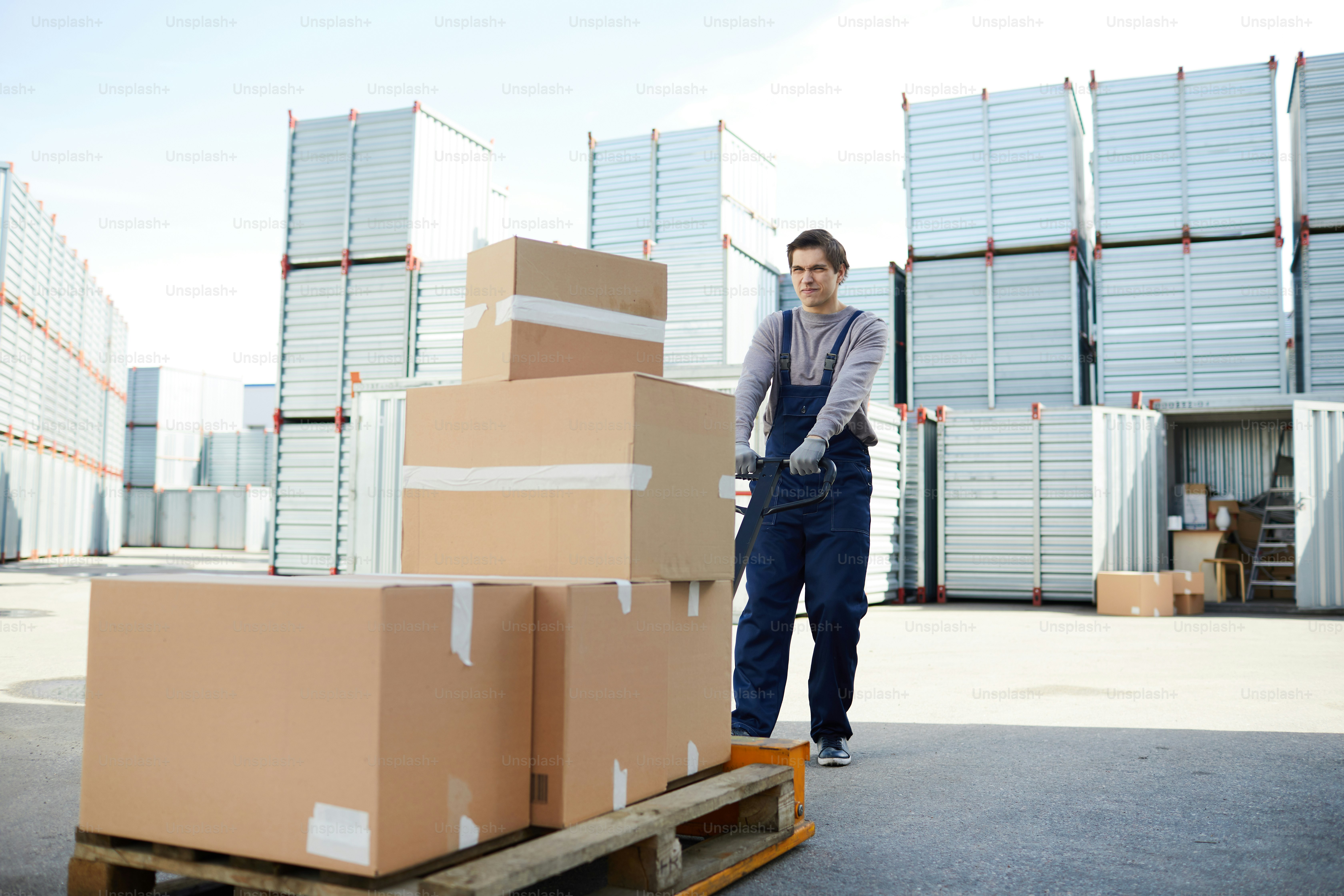 Frowning young man pulling heavy pullet jack with stack of boxes while ...