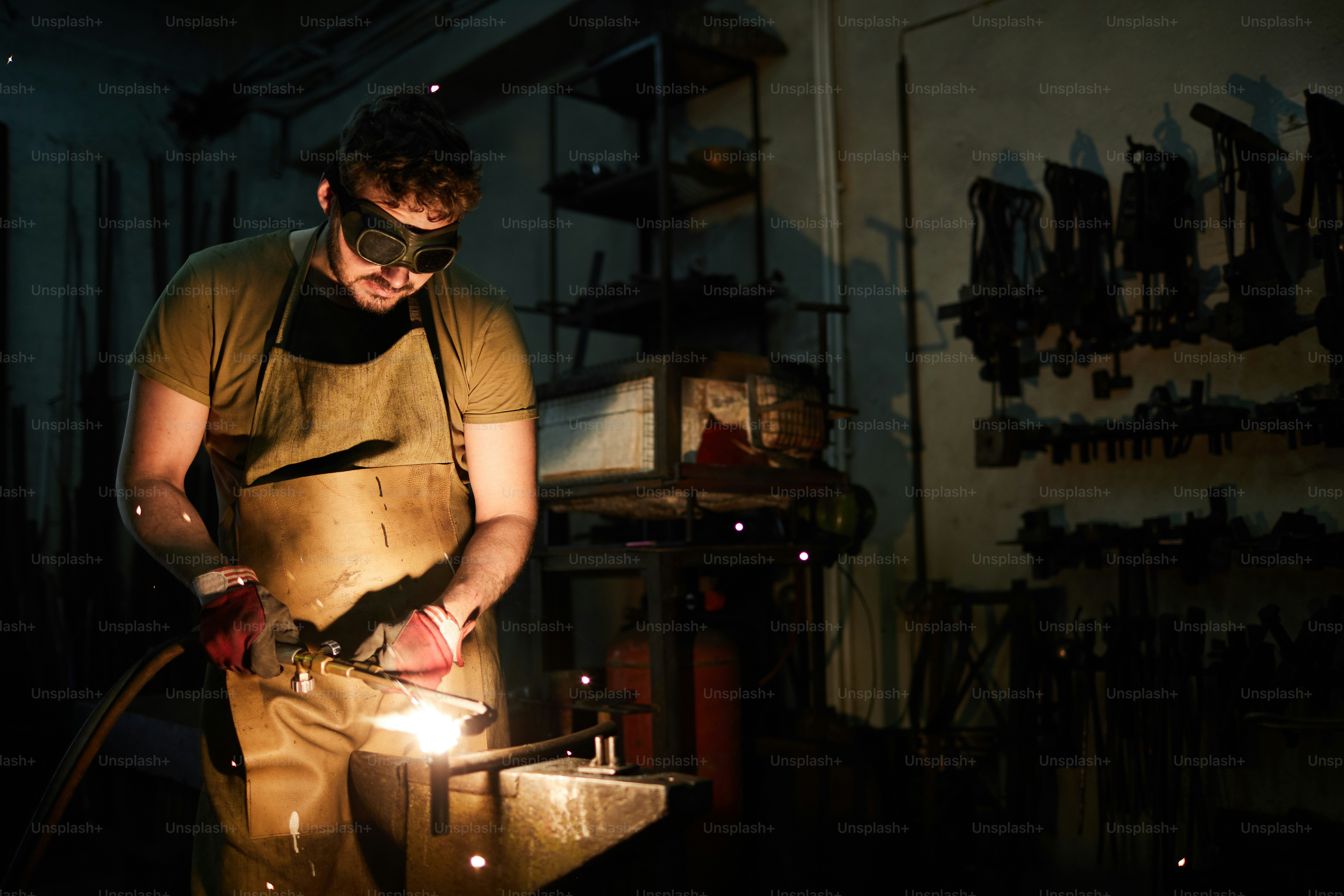 Young blacksmith in protective eyeglasses and apron standing by anvil ...