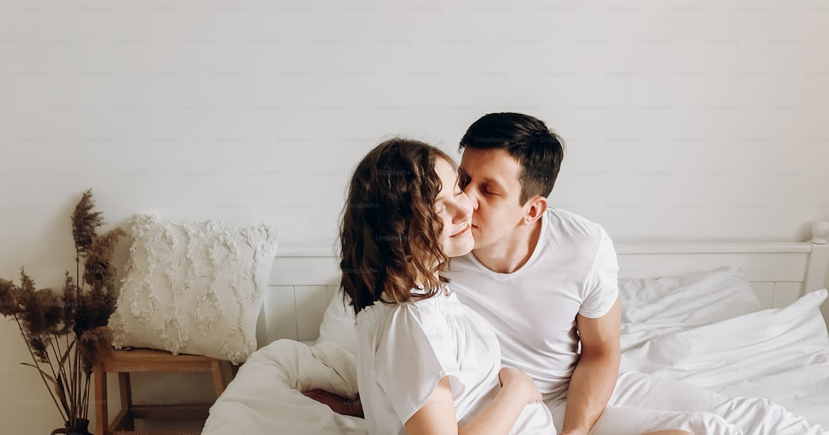 행복한 젊은 남편은 웃고 있는 임신한 아내에게 키스하고 하얀 침대에서 배꼽 범프를 껴안고 있다 흰색의 세련된 임신 한 커플은 집에서 배를 잡고 있습니다 다산 개념입니다