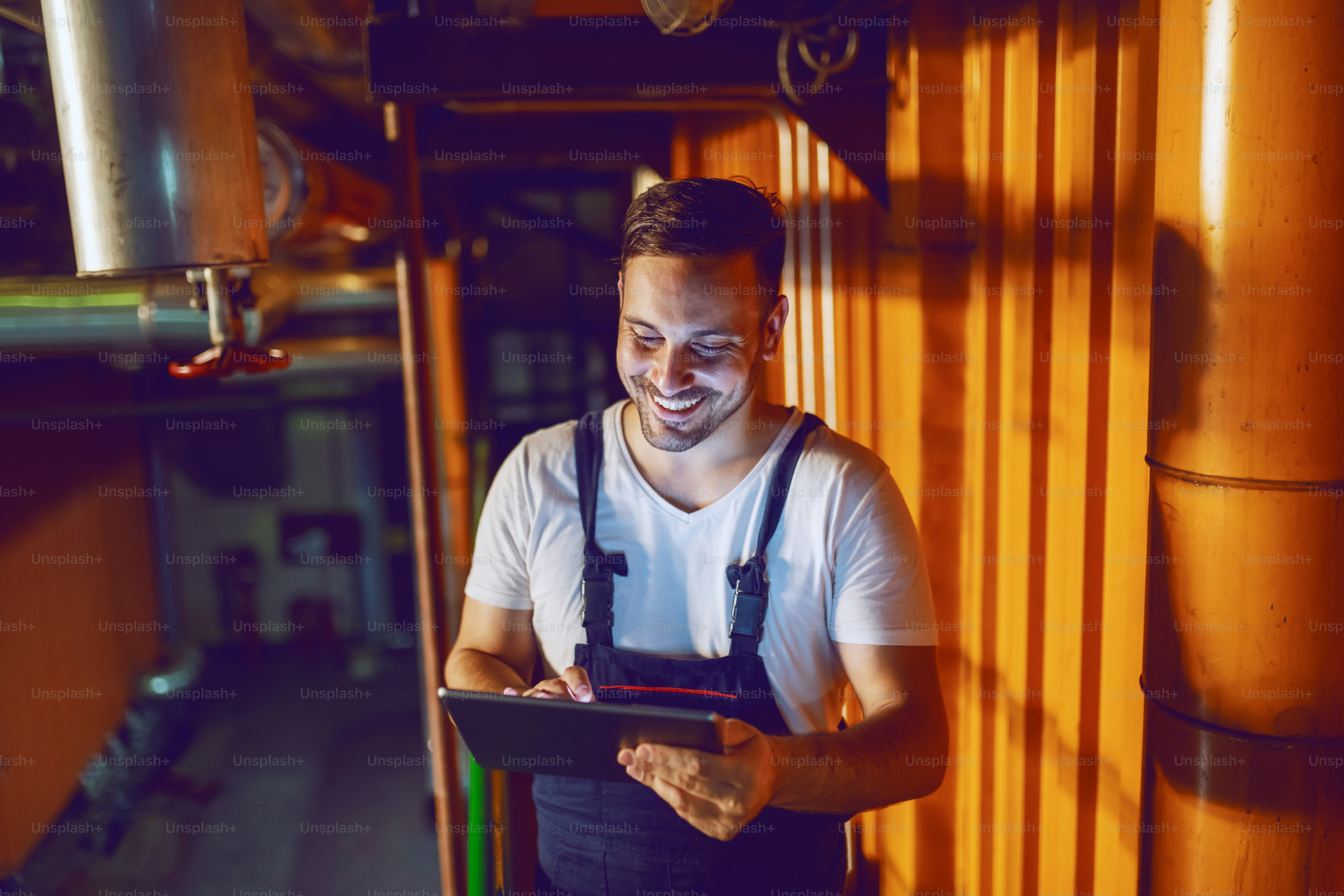 Smiling hardworking plant worker in coveralls standing and using tablet ...