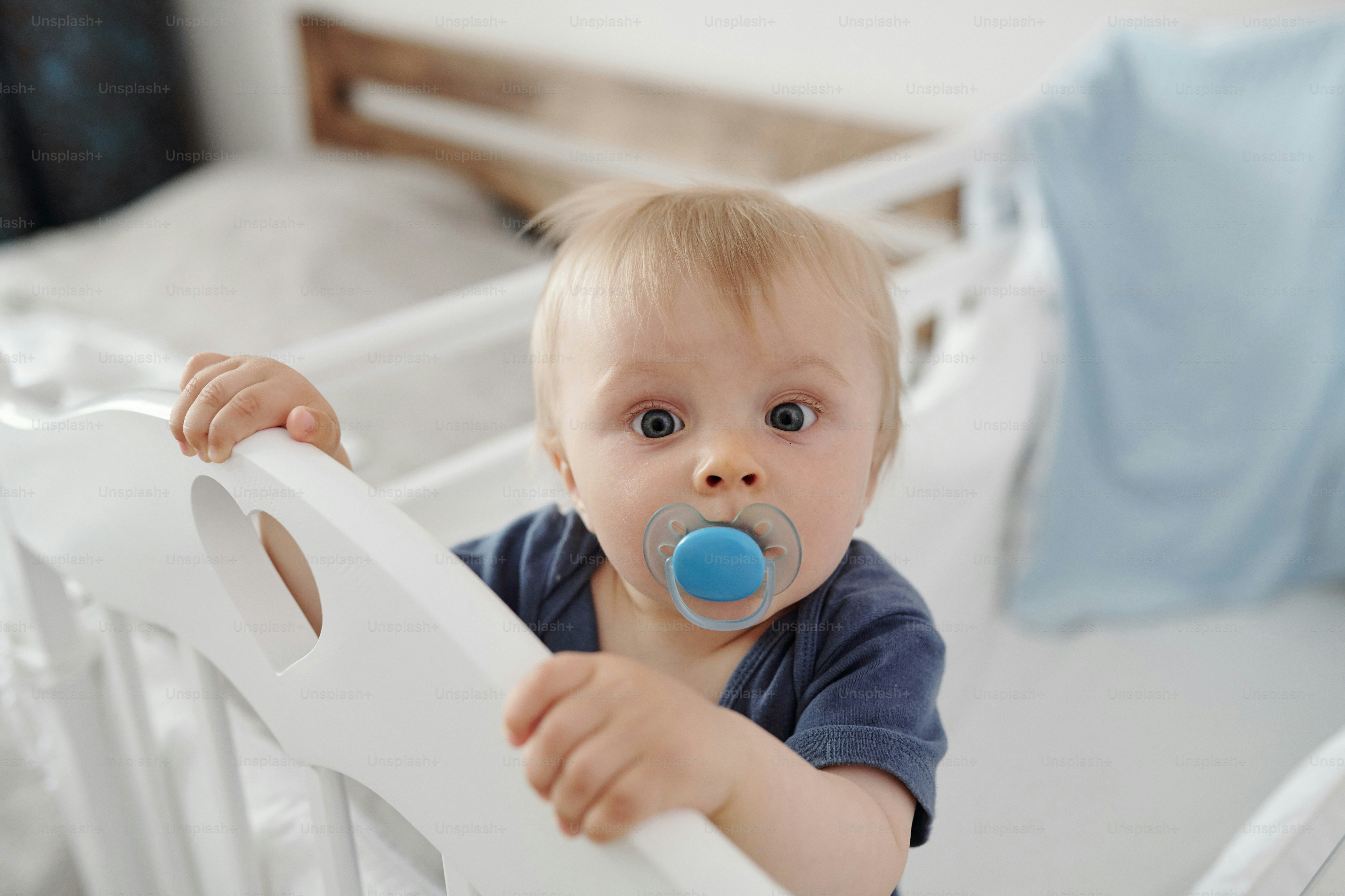 Portrait of curious blond-haired baby with dummy holding barred side of ...