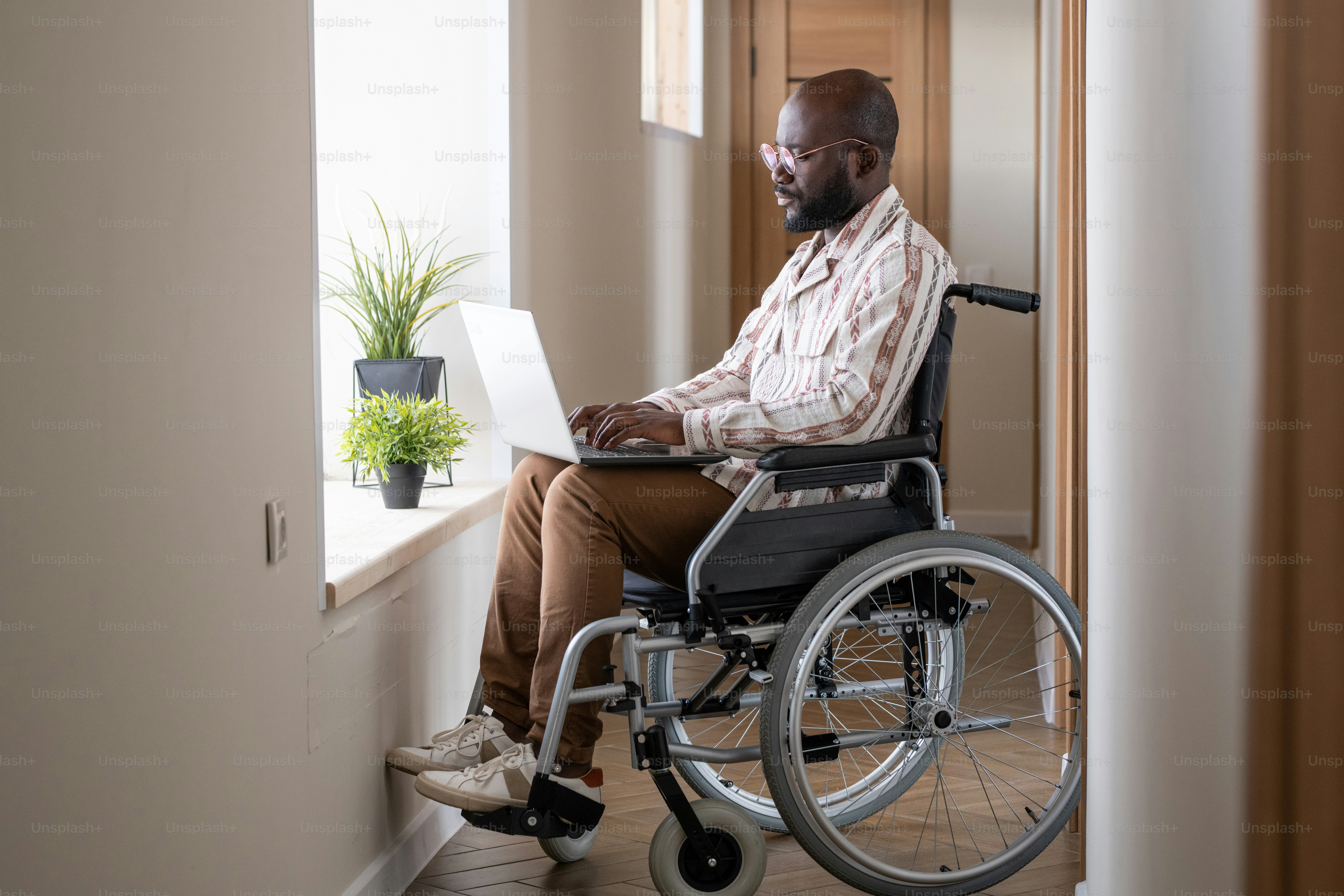 Young serious businessman or freelancer in wheelchair sitting by table in living room and ...