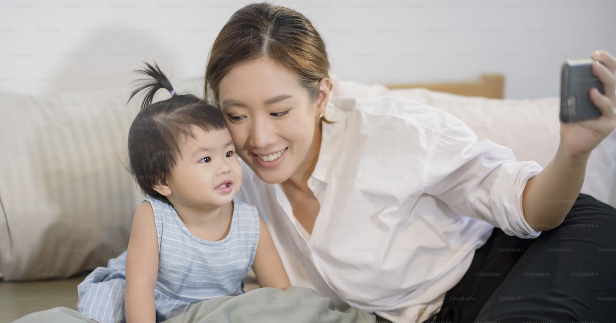아시아 어머니와 그녀의 아기 딸은 침대 가족 가정 안전 부모 기술 개념에서 아버지에게 셀카 또는 화상 통화를 하고