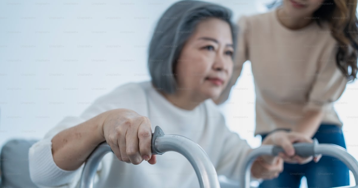 아시아의 어린 딸은 집에서 보행기와 함께 걷는 늙은 장애인 여성을 지원합니다 아름 다운 소녀는 집에서 거실에서 물리 치료를