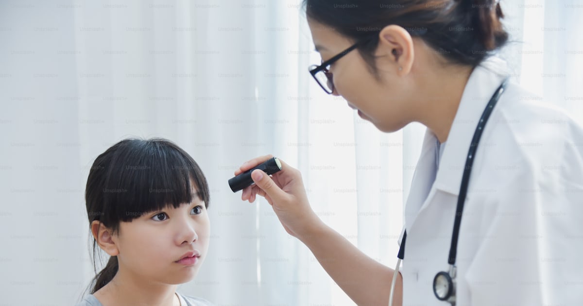 젊은 아시아 여성 의사는 의료 사무실에서 손전등으로 어린 소녀 환자의 눈을 확인하는 안경을 착용합니다 사진 Unsplash