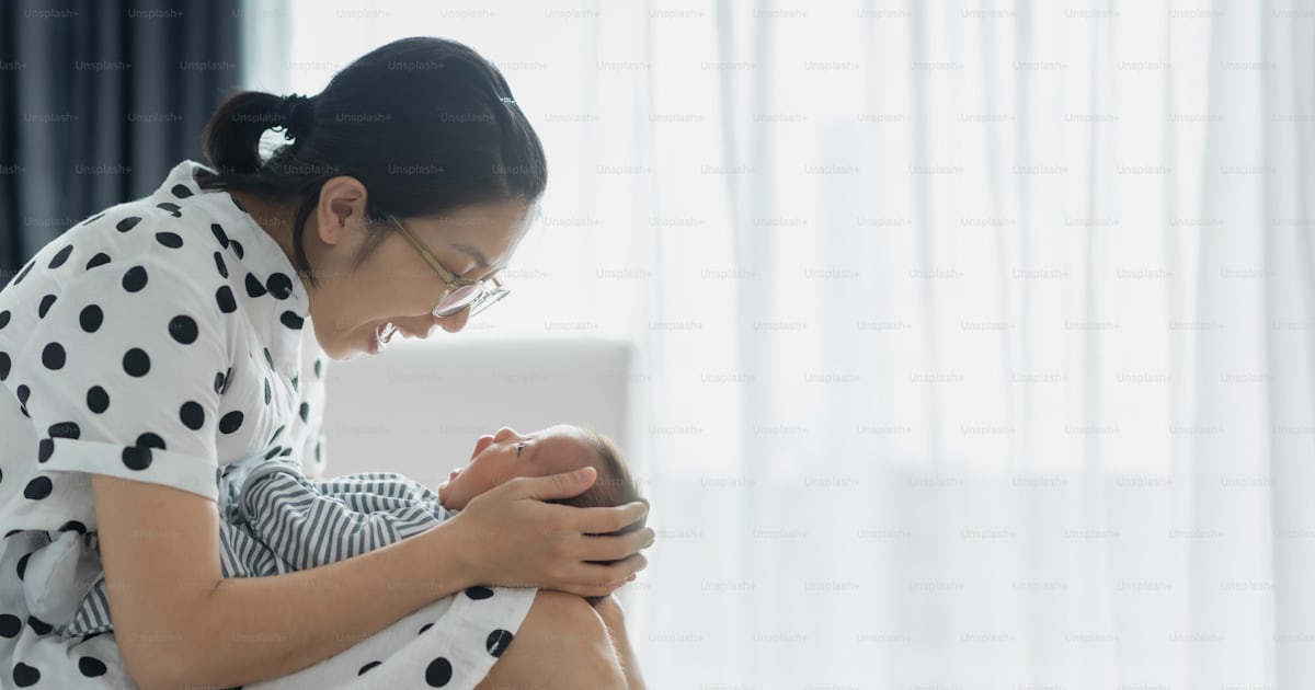 행복한 아시아 젊은 엄마는 침대에 앉아 작은 유아와 놀고 함께 웃고 있다 사랑하는 여성은 아들을 손에 껴안고 즐긴다 엄마와