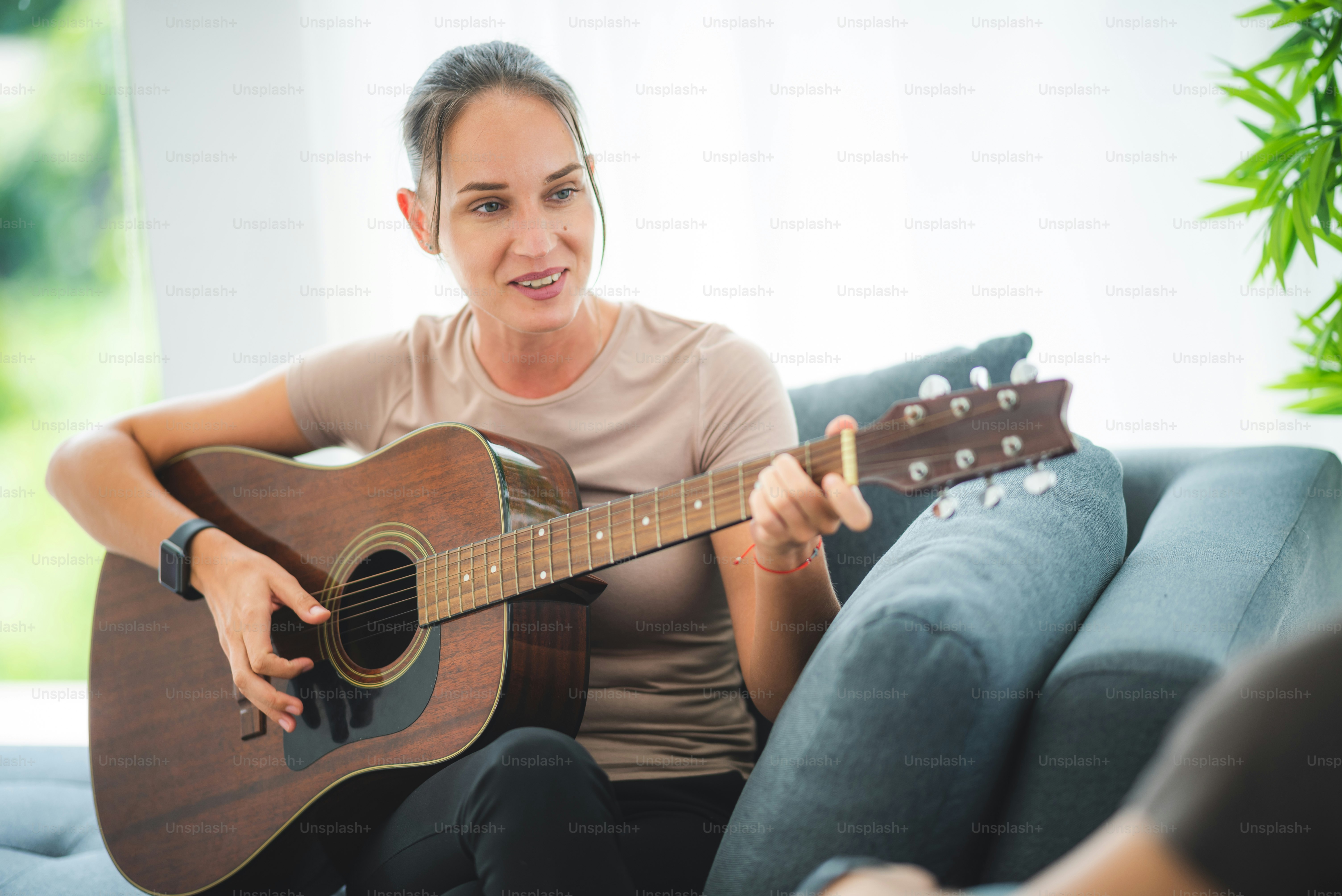 Una hermosa mujer tocando una guitarra acústica mientras está sentada en un sofá con fondo ...