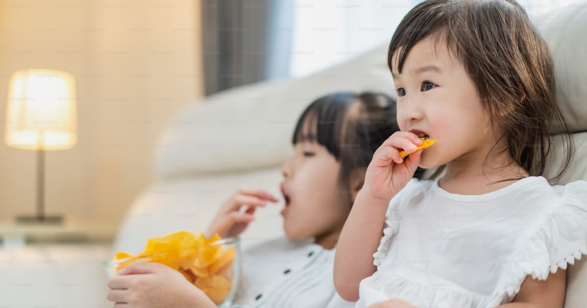 아시아 배고픈 어린 소녀 형제 자매 손으로 간식을 입에 넣습니다 집에서 소파에서 쉬고 있는 두 명의 달콤한 미취학 아동은