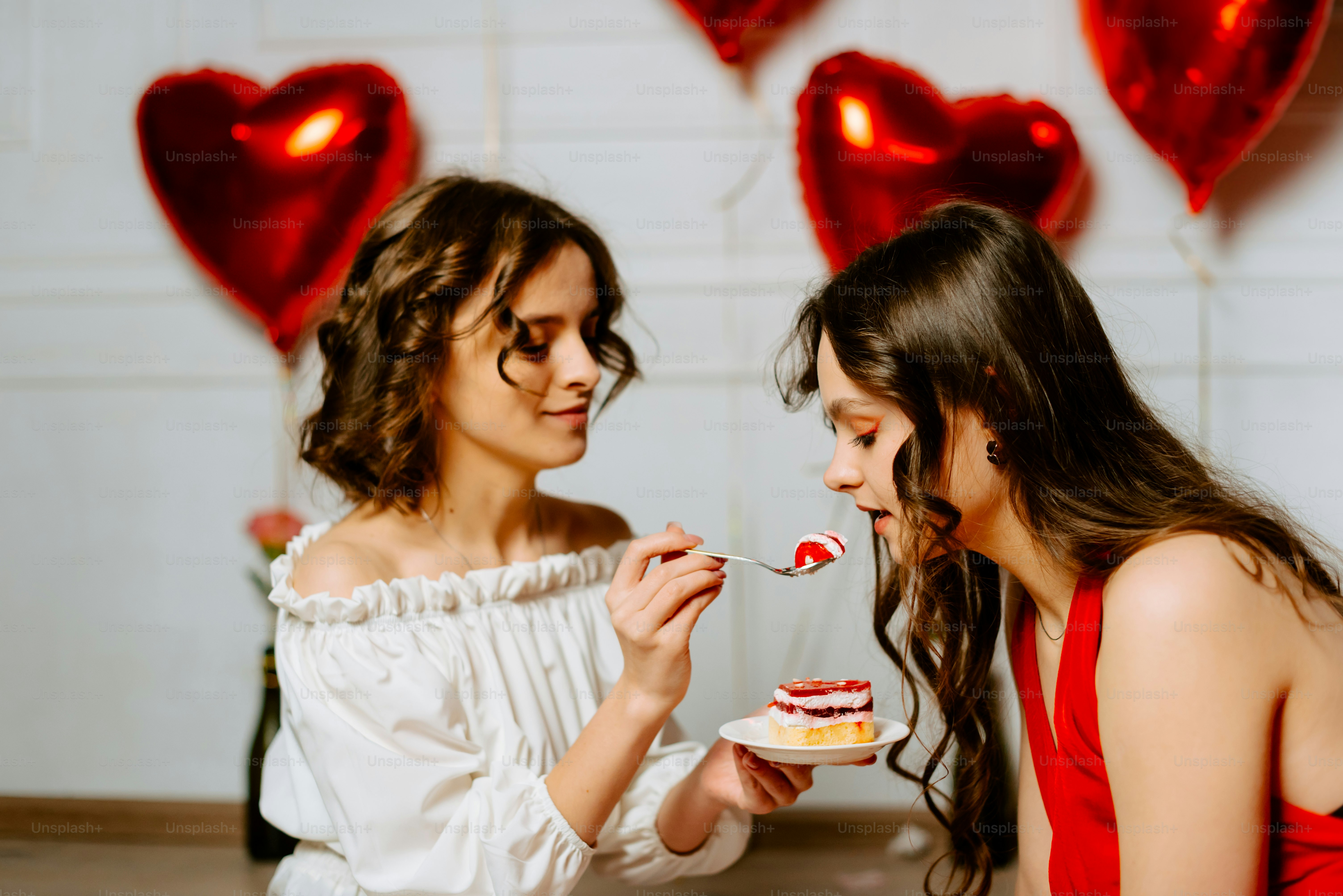 Two women are eating cake together in front of heart shaped balloons ...