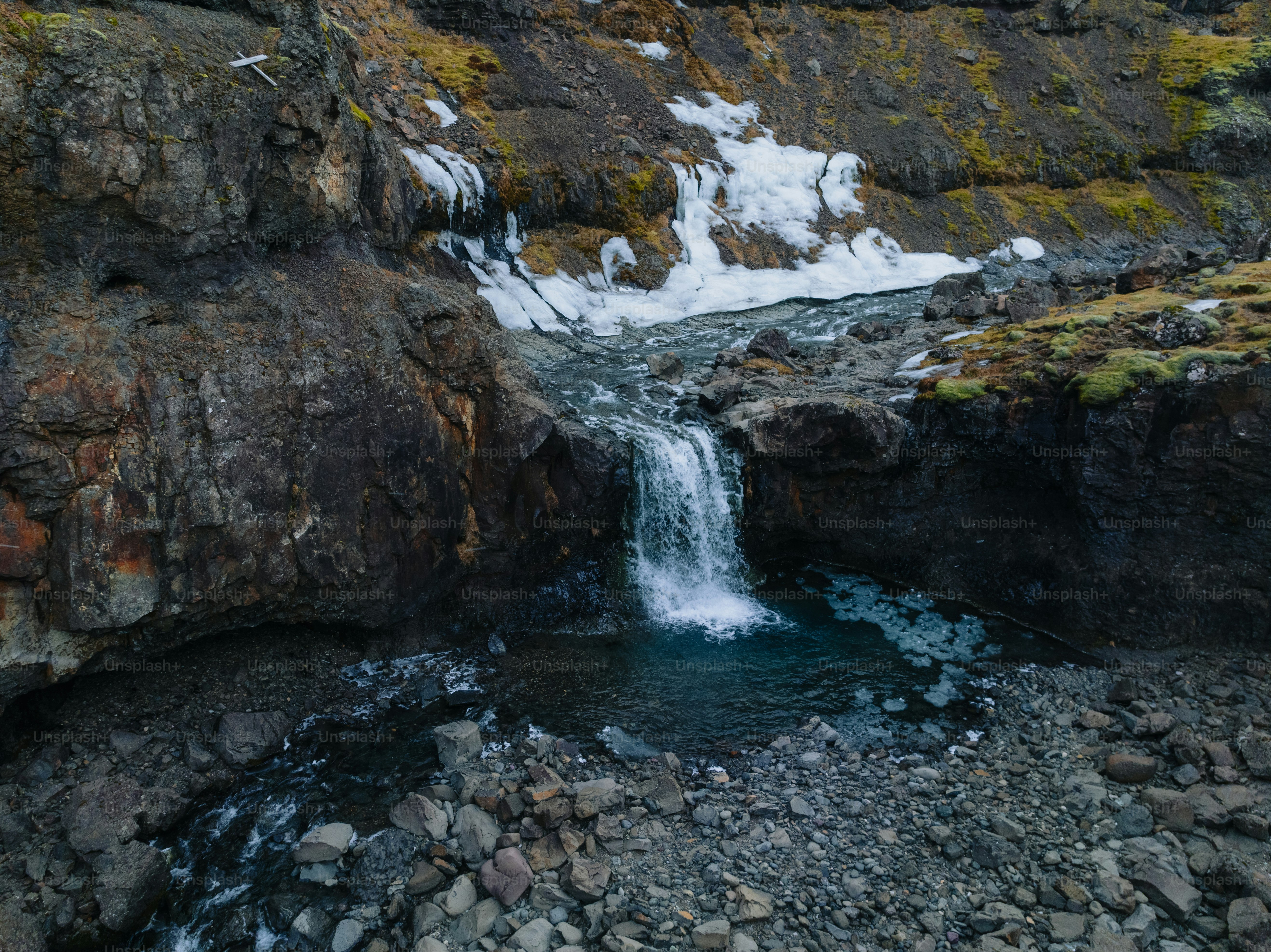 A small waterfall in the middle of a rocky area photo – Patches of snow ...