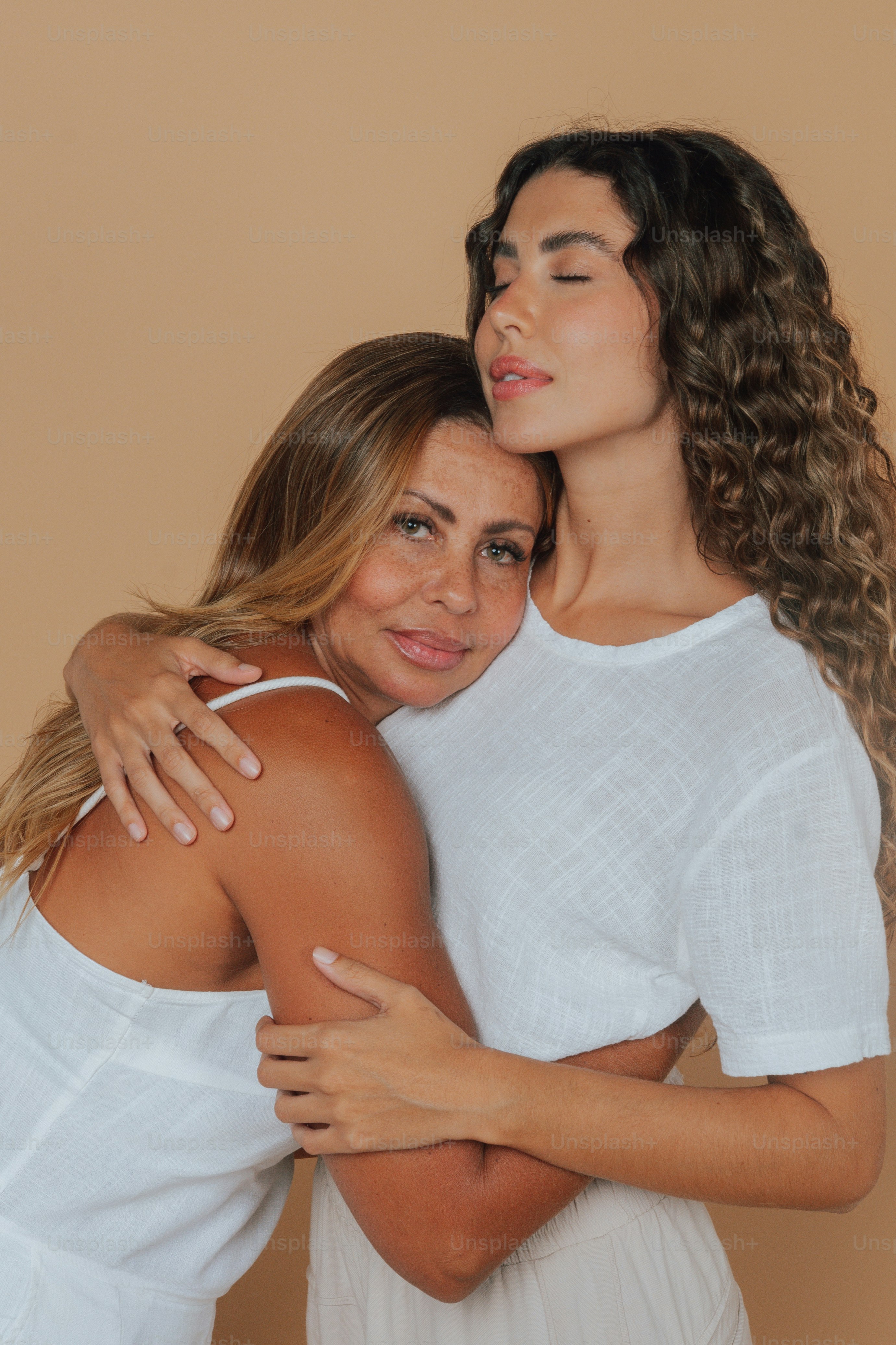 Two beautiful women hugging each other in front of a beige background ...
