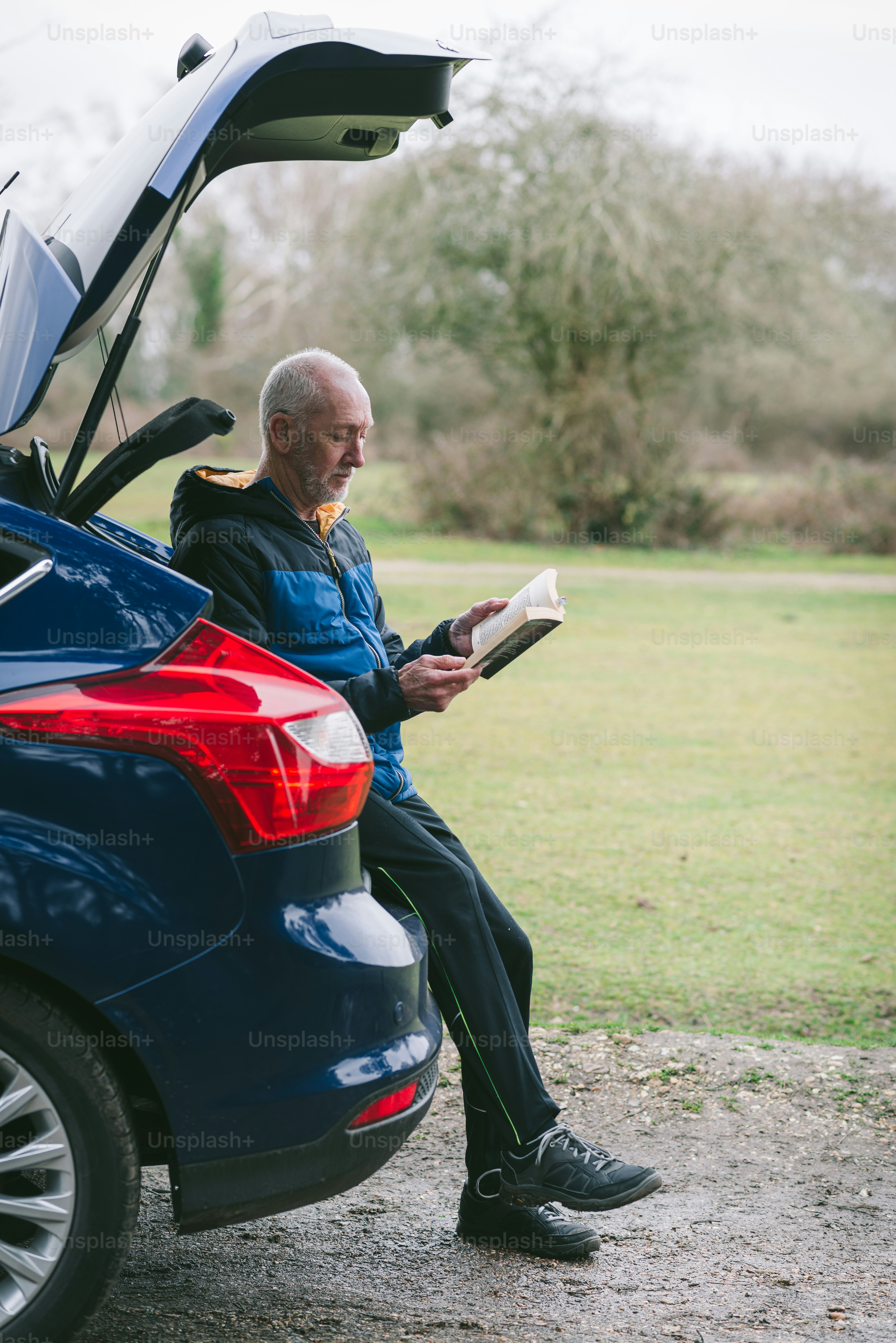 A man sitting on the trunk of a car reading a book photo – Reading a ...