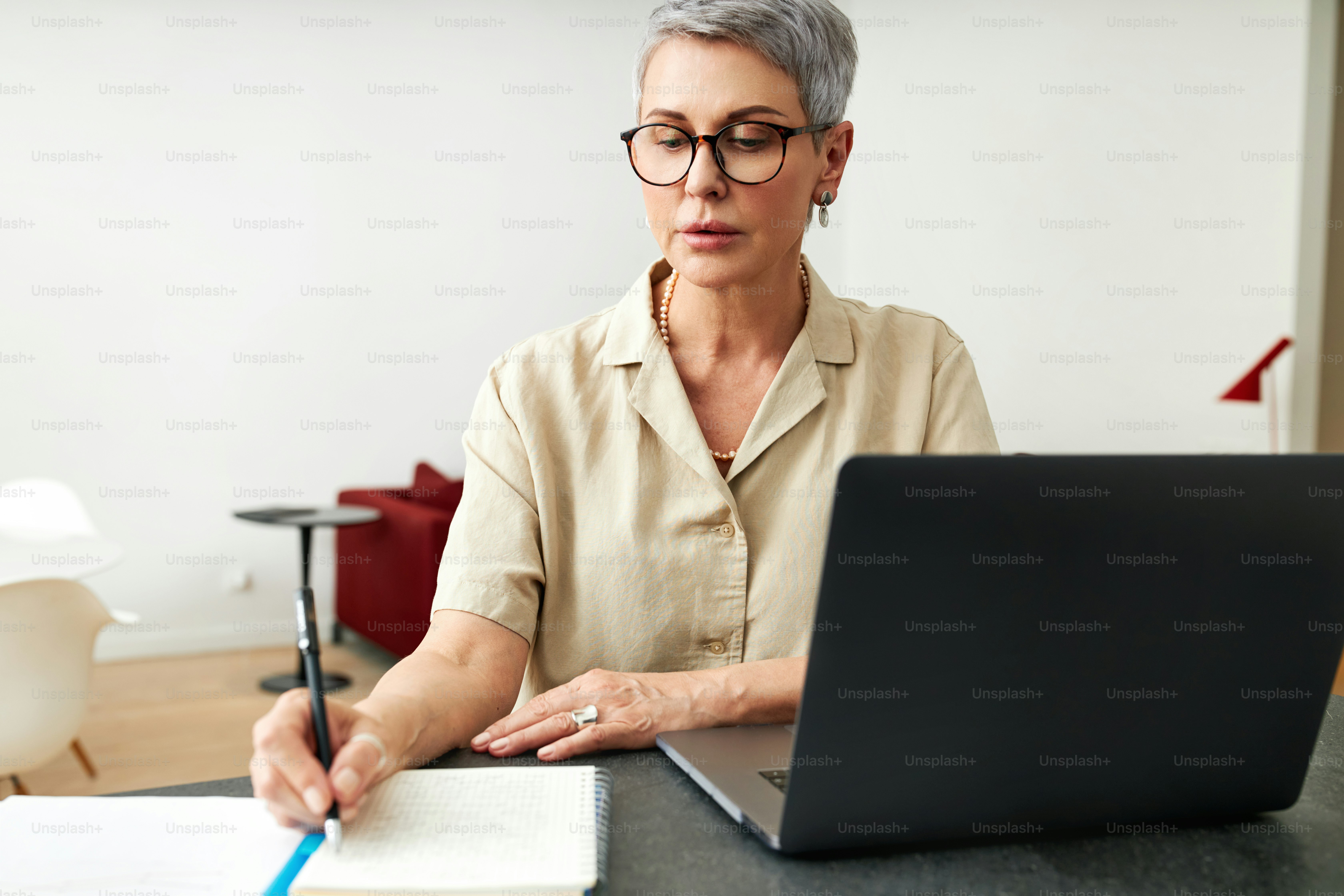 Mature woman writing on a note pad while using laptop computer at home ...