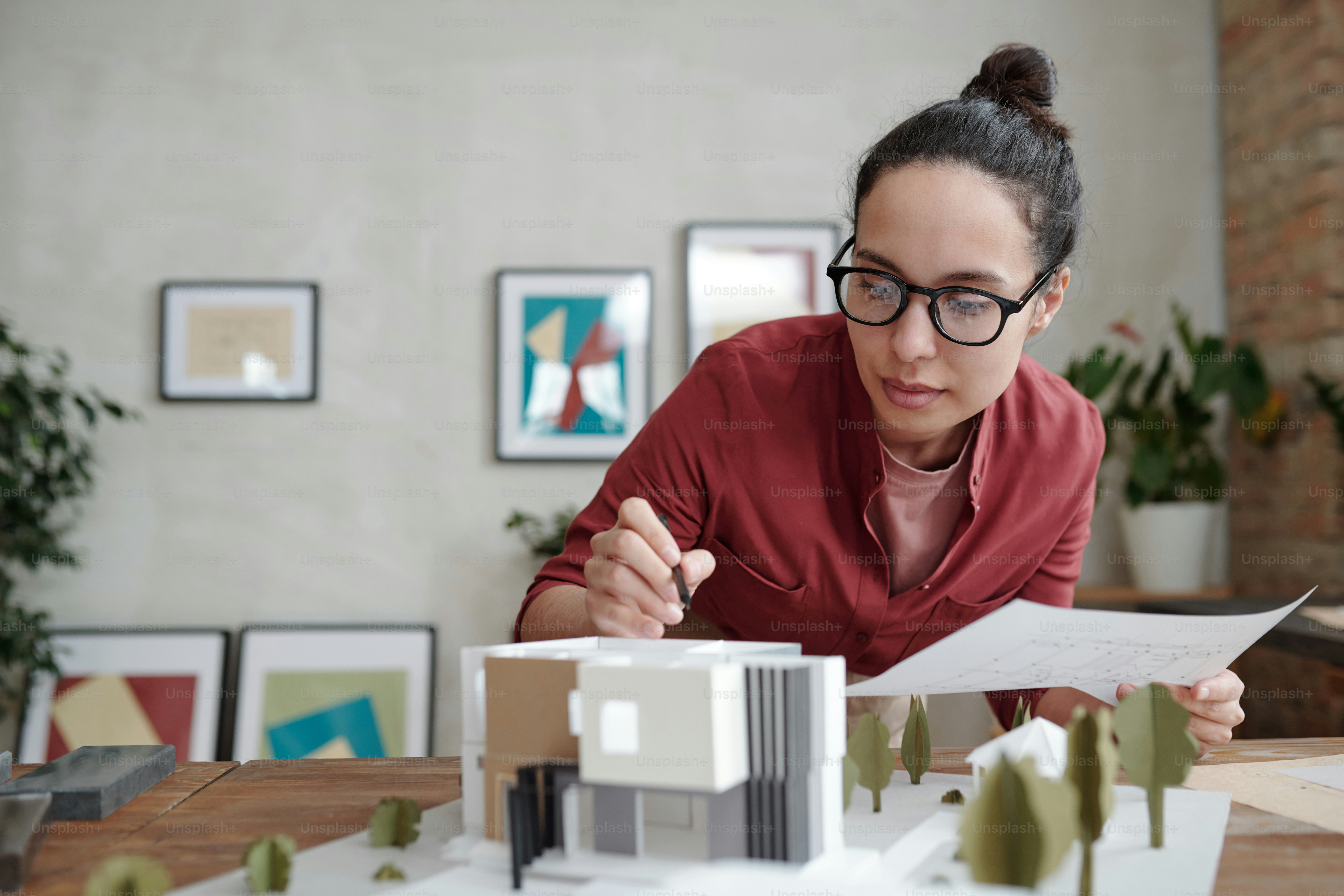 Young elegant female architect with paper and pen bending over model of ...