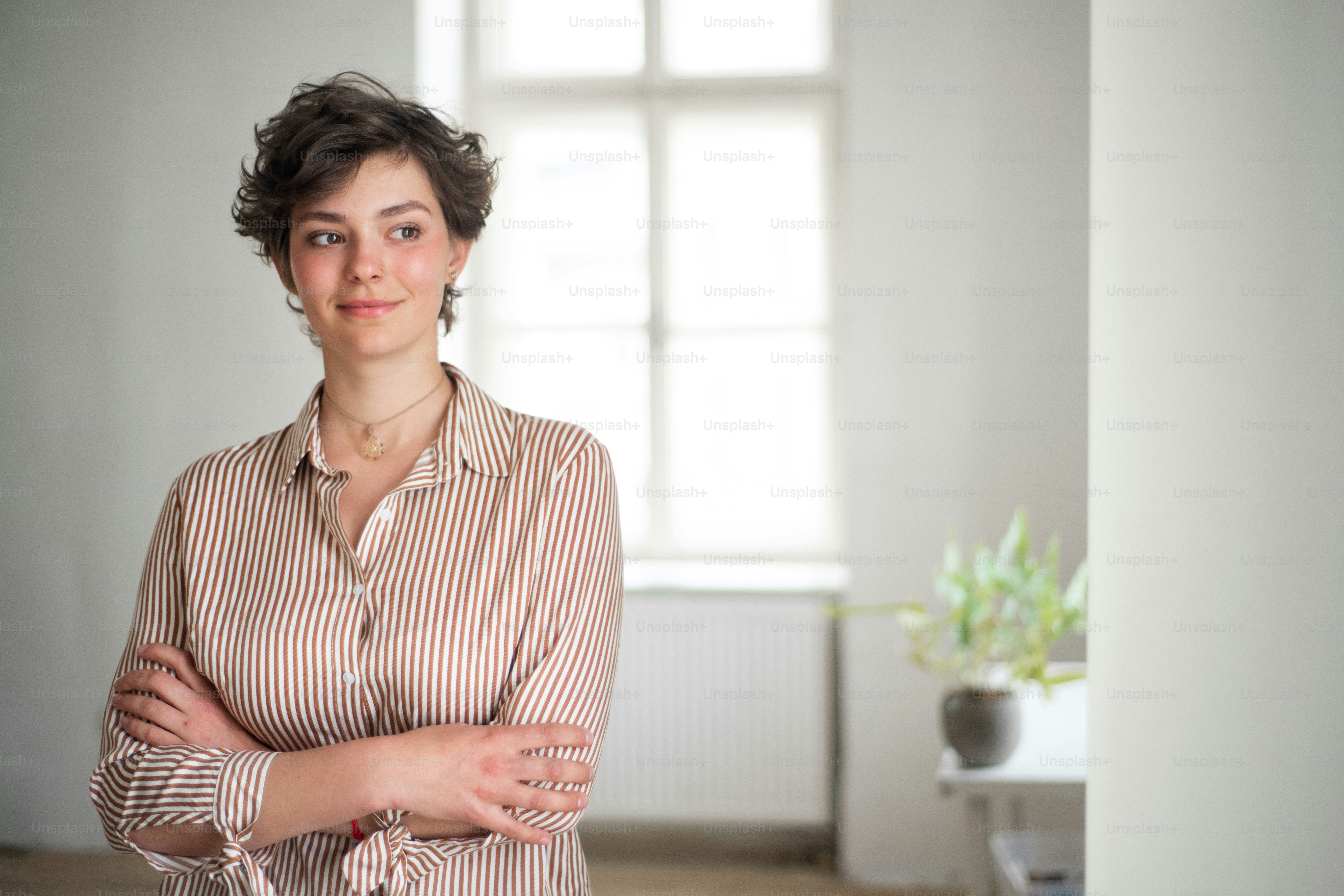 A happy young woman student standing in front of window in office ...