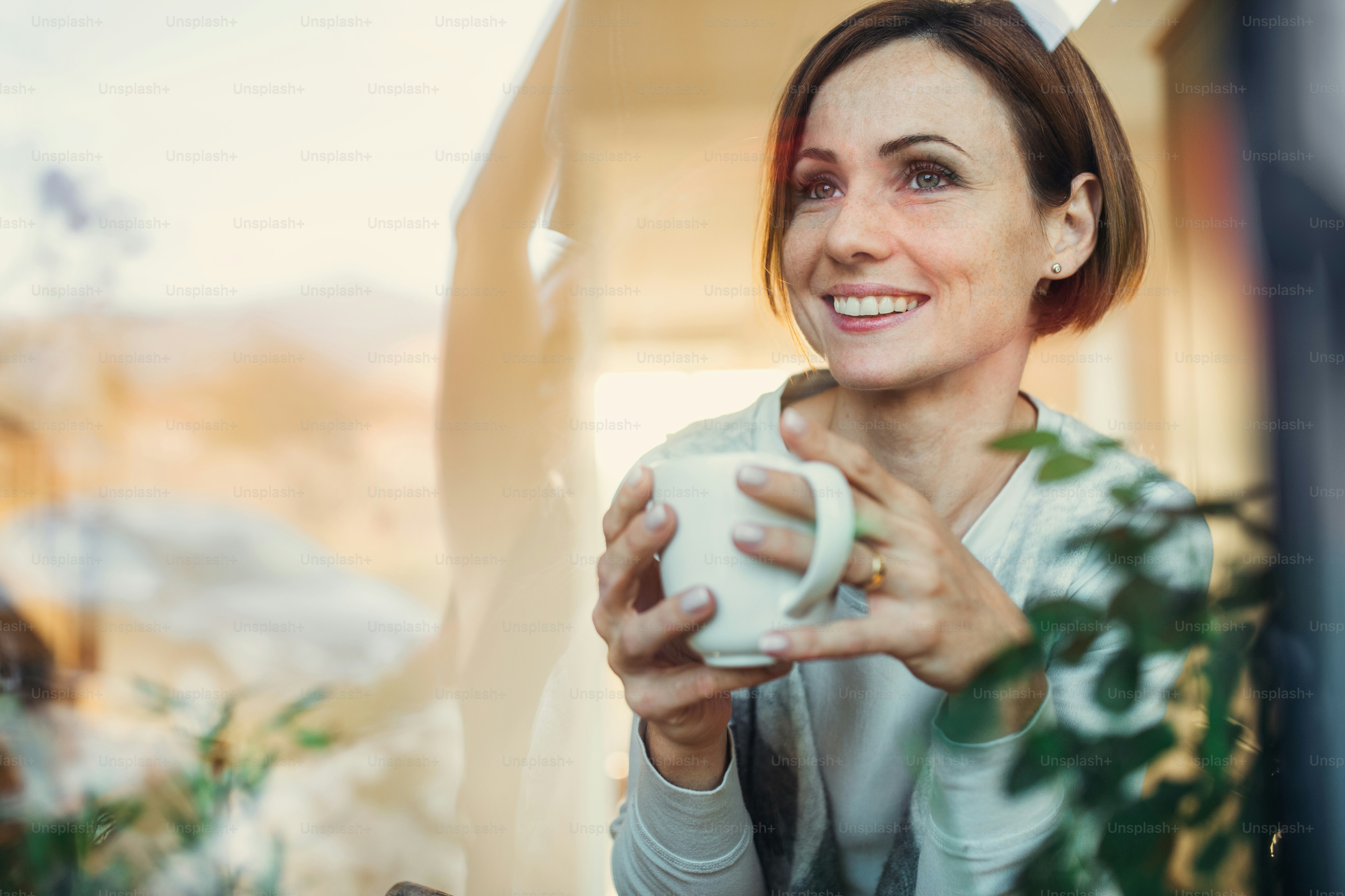 A midsection of young woman with cup of coffee looking out of a window ...