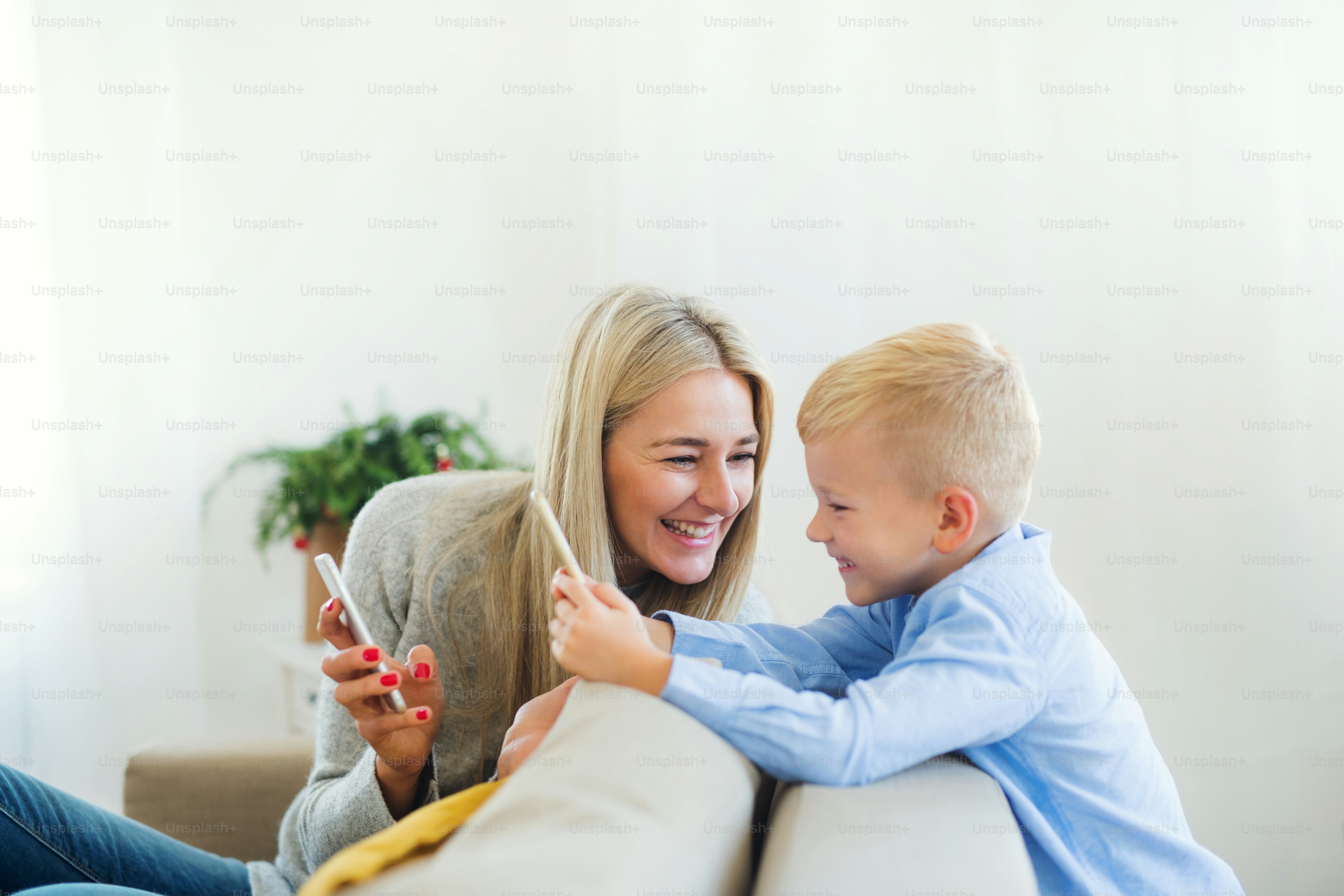 A mother and small boy with smartphones sitting on a sofa at home at Christmas time, playing ...
