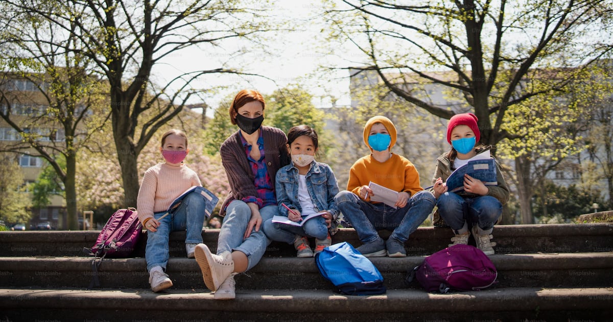 都市公園の屋外に座って、グループ教育とコロナウイルスのコンセプトを学ぶ小さな子供を持つ教師。の写真 Unsplashの人の画像