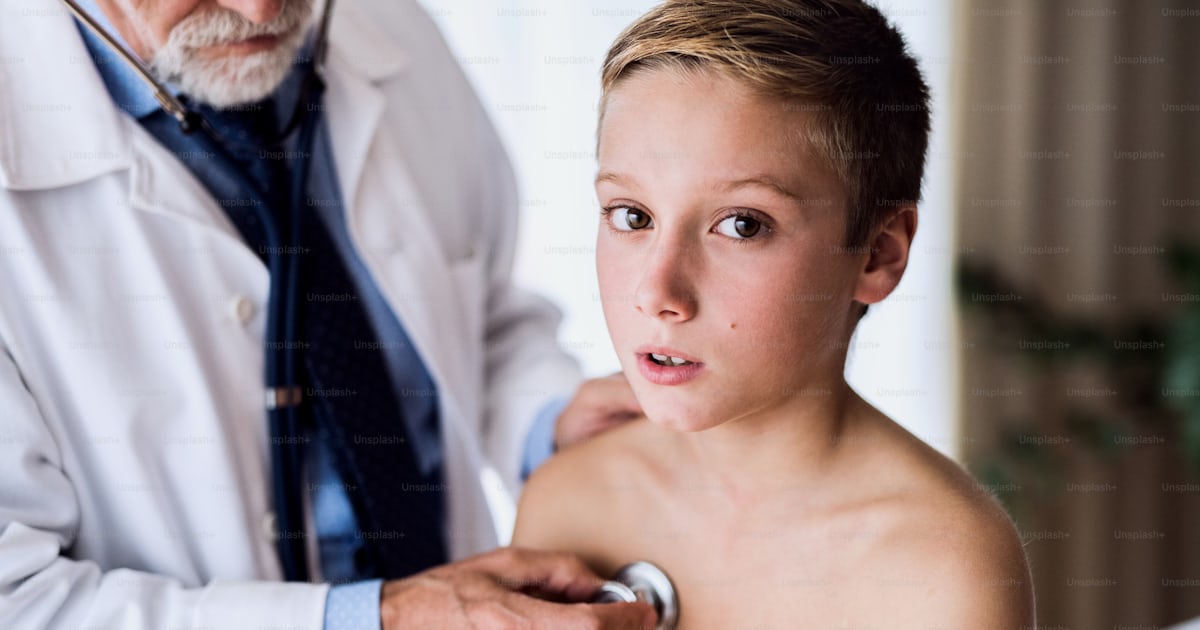 彼のオフィスで聴診器で小さな男の子を診察している認識できない年配の男性医師。の写真 Unsplashの調べるの画像