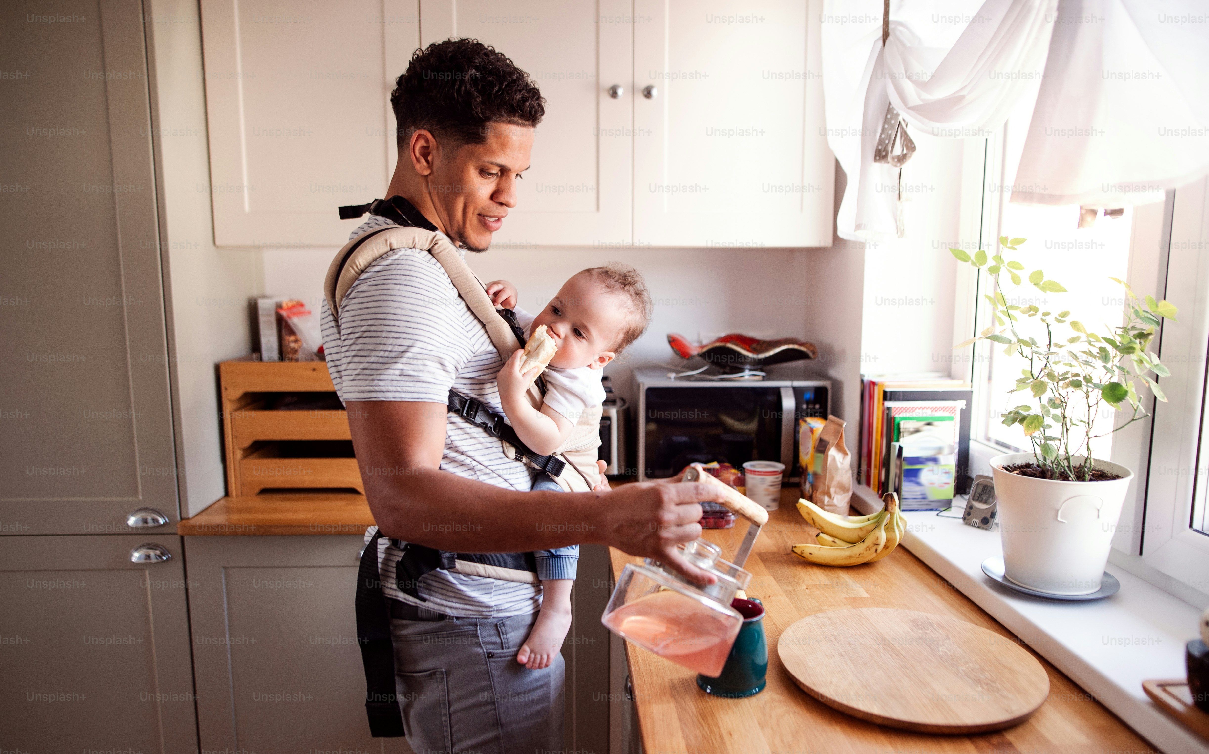 A father with small toddler son in carrier in kitchen indoors at home ...
