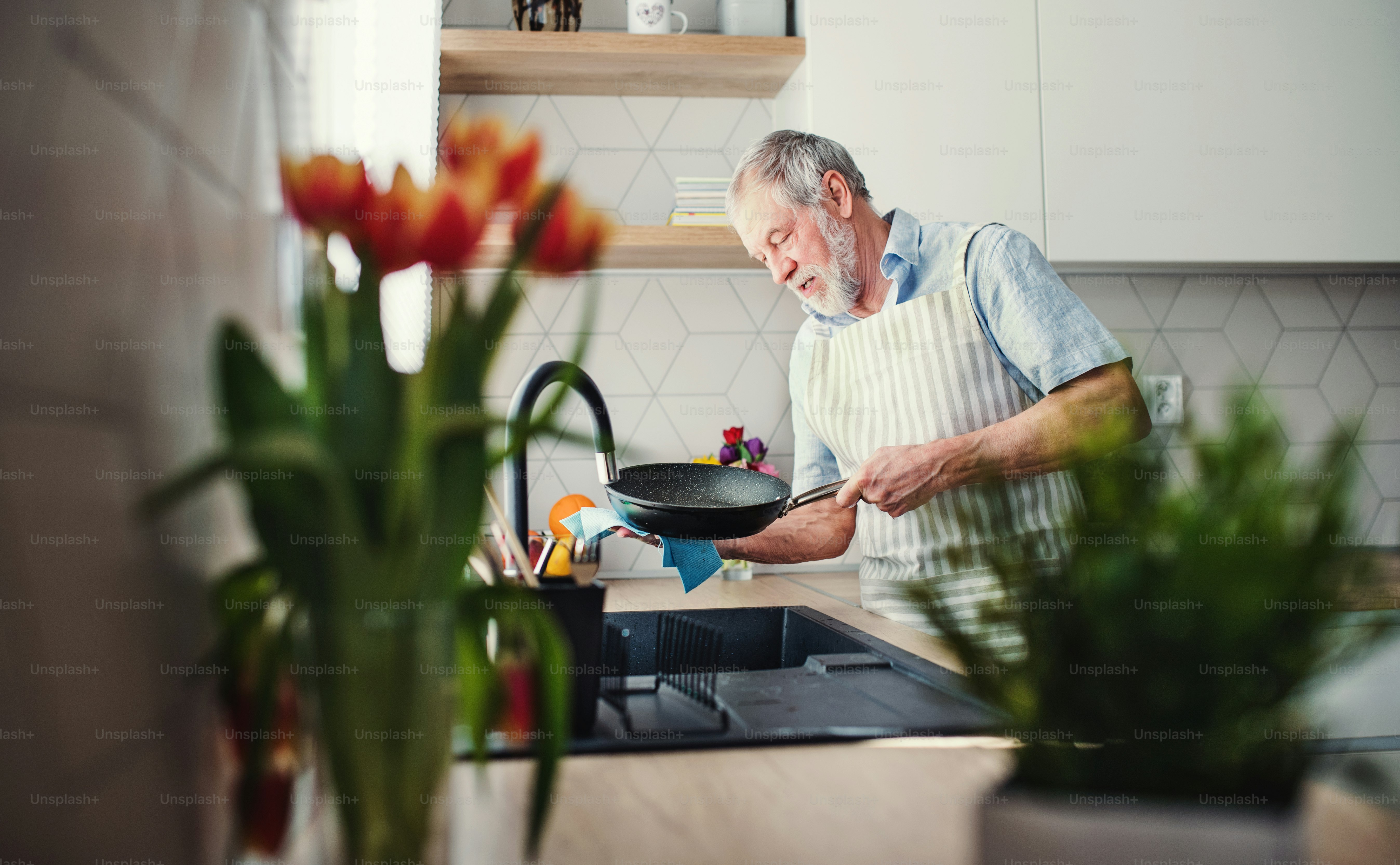 A senior man indoors in kitchen at home, washing up a pan after cooking ...