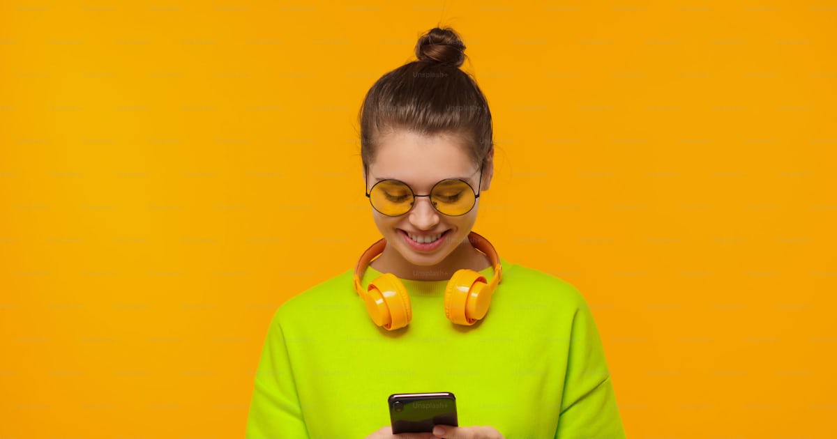 黄色い背景に笑顔でスマートフォンの画面を見る首にヘッドフォンを持つ若い女性の写真 幸福 Unsplashの写真