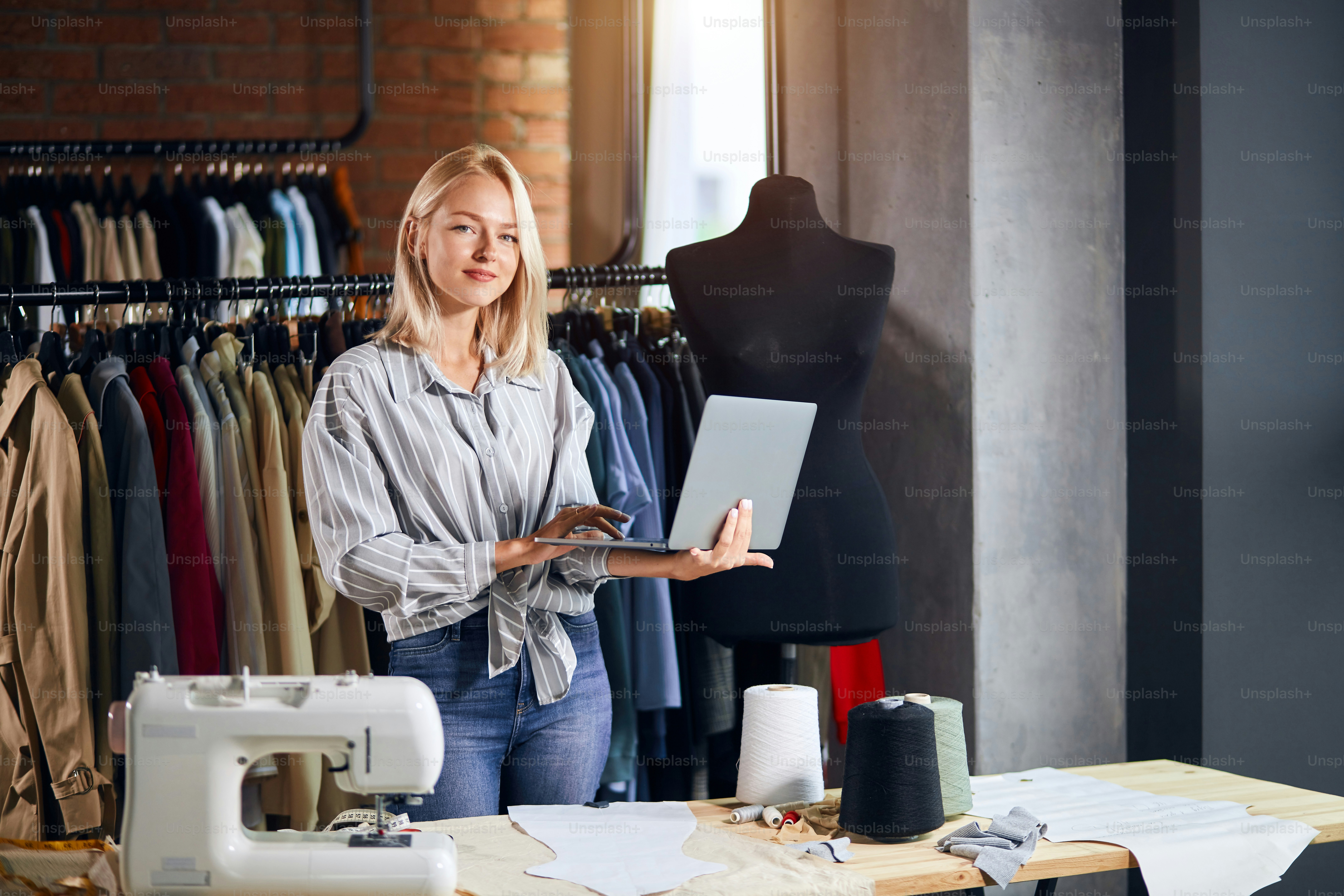 Awesome young female dressmaker holding laptop, looking at the camera ...