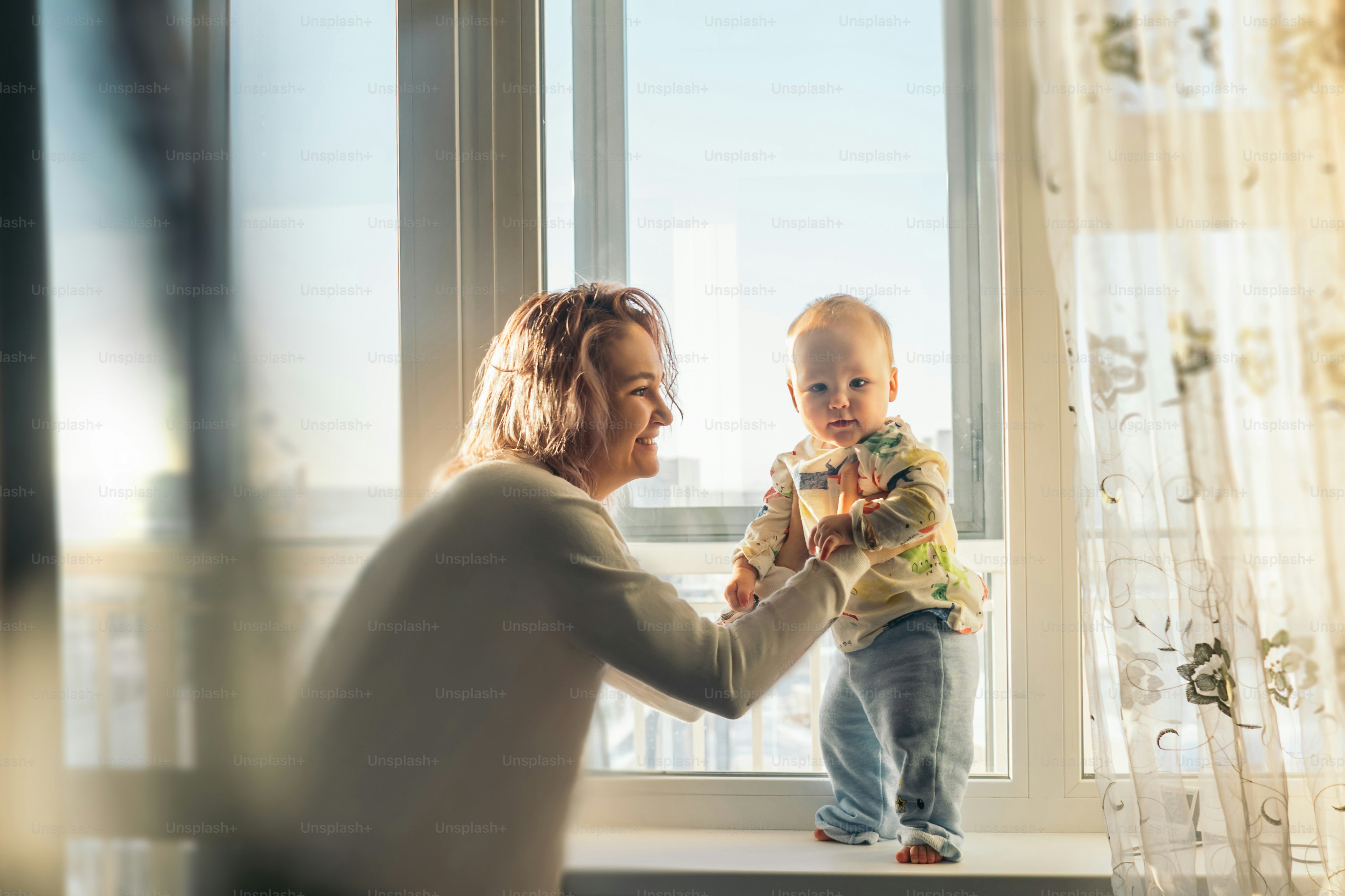 Happy young mother with cute baby boy on windowsill. At home near ...