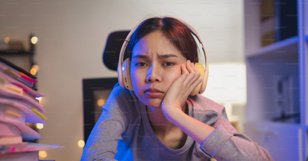 스트레스가 많은 젊은 아시아 여성은 헤드셋을 끼고 의자에 앉아 탁자 위에 서류 더미를 놓고 밤에 컴퓨터 화면을 보고 있다 사진