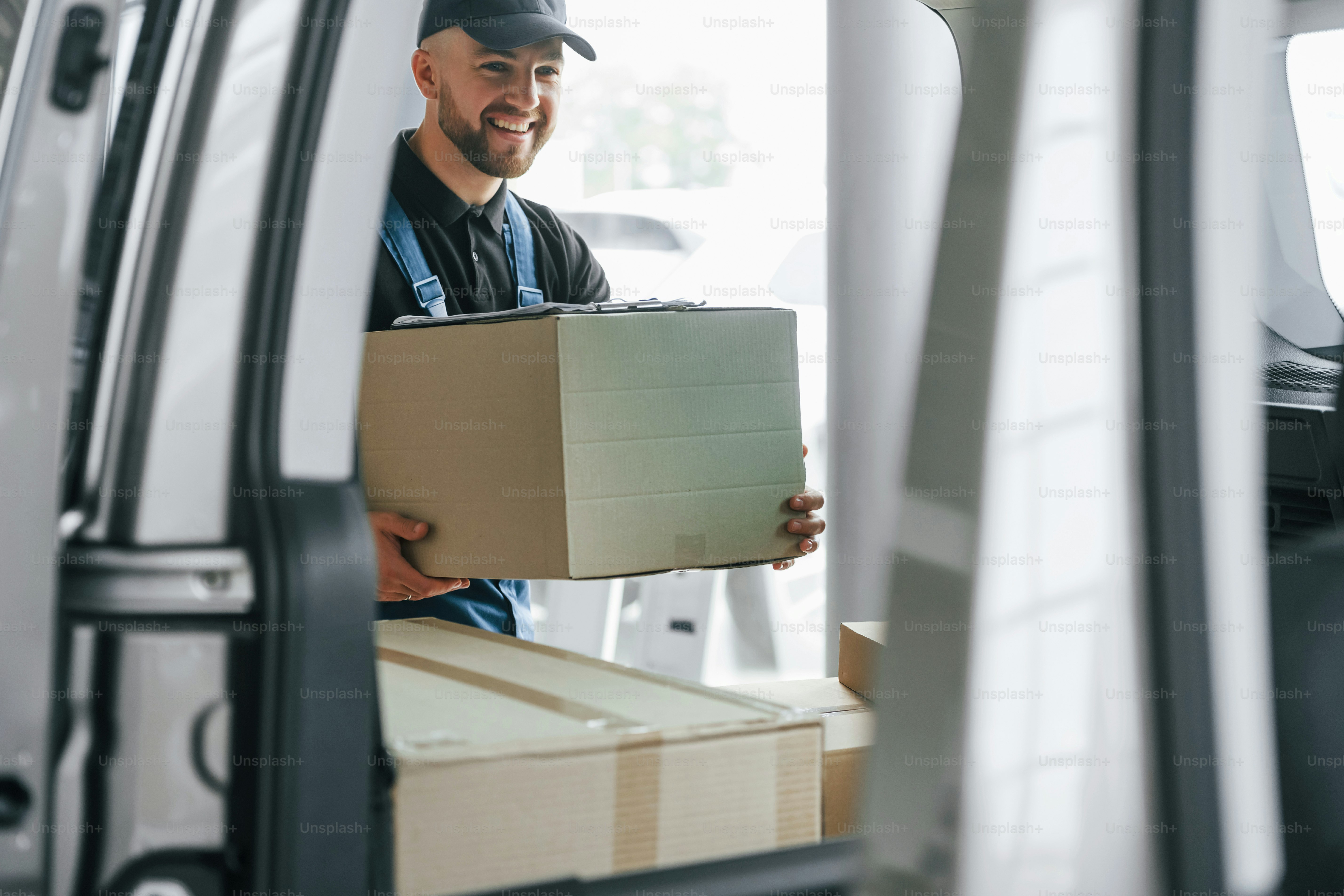 Cheerful worker. Delivery man in uniform is indoors with car and with ...