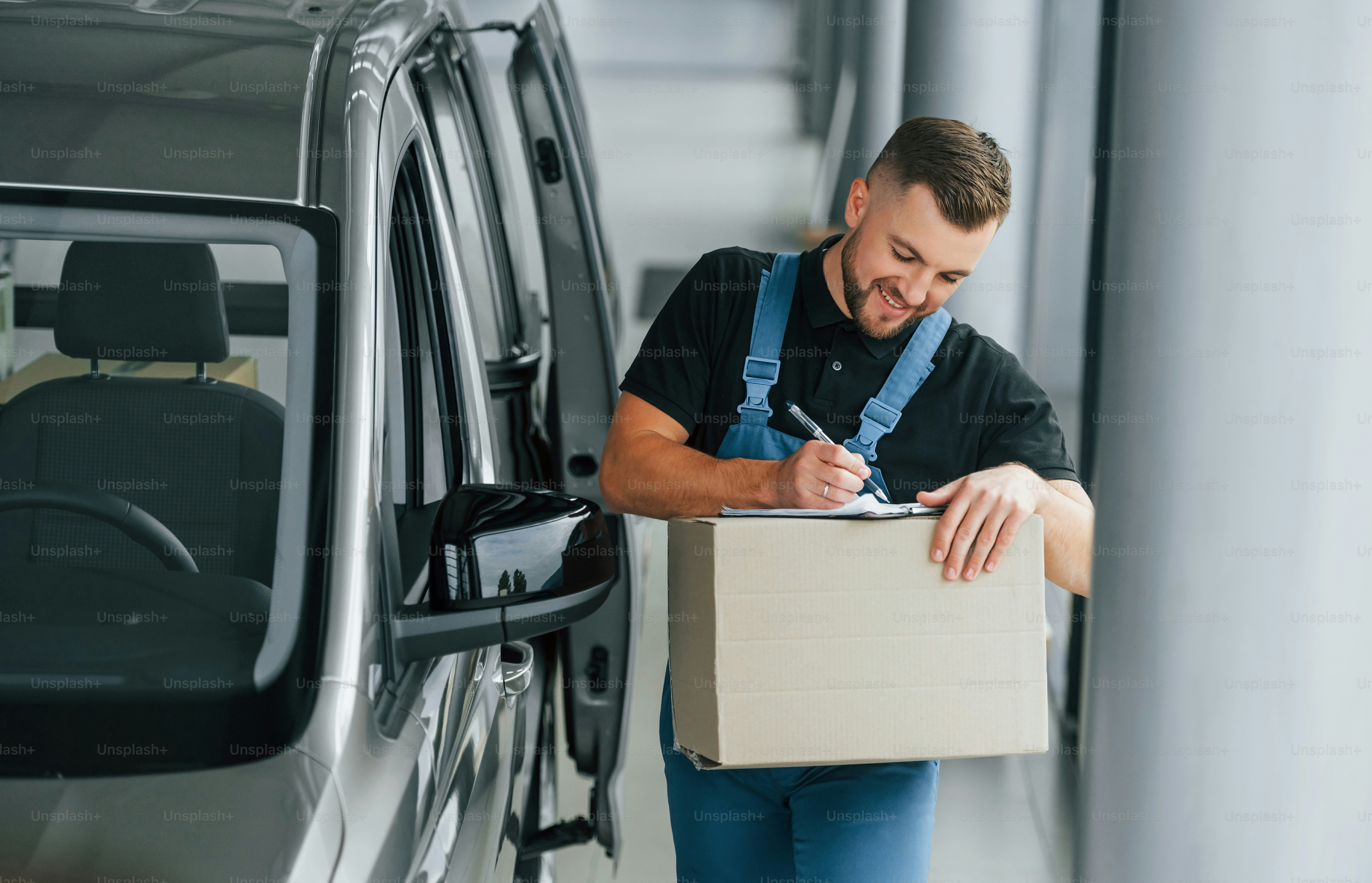 Cheerful worker. Delivery man in uniform is indoors with car and with ...