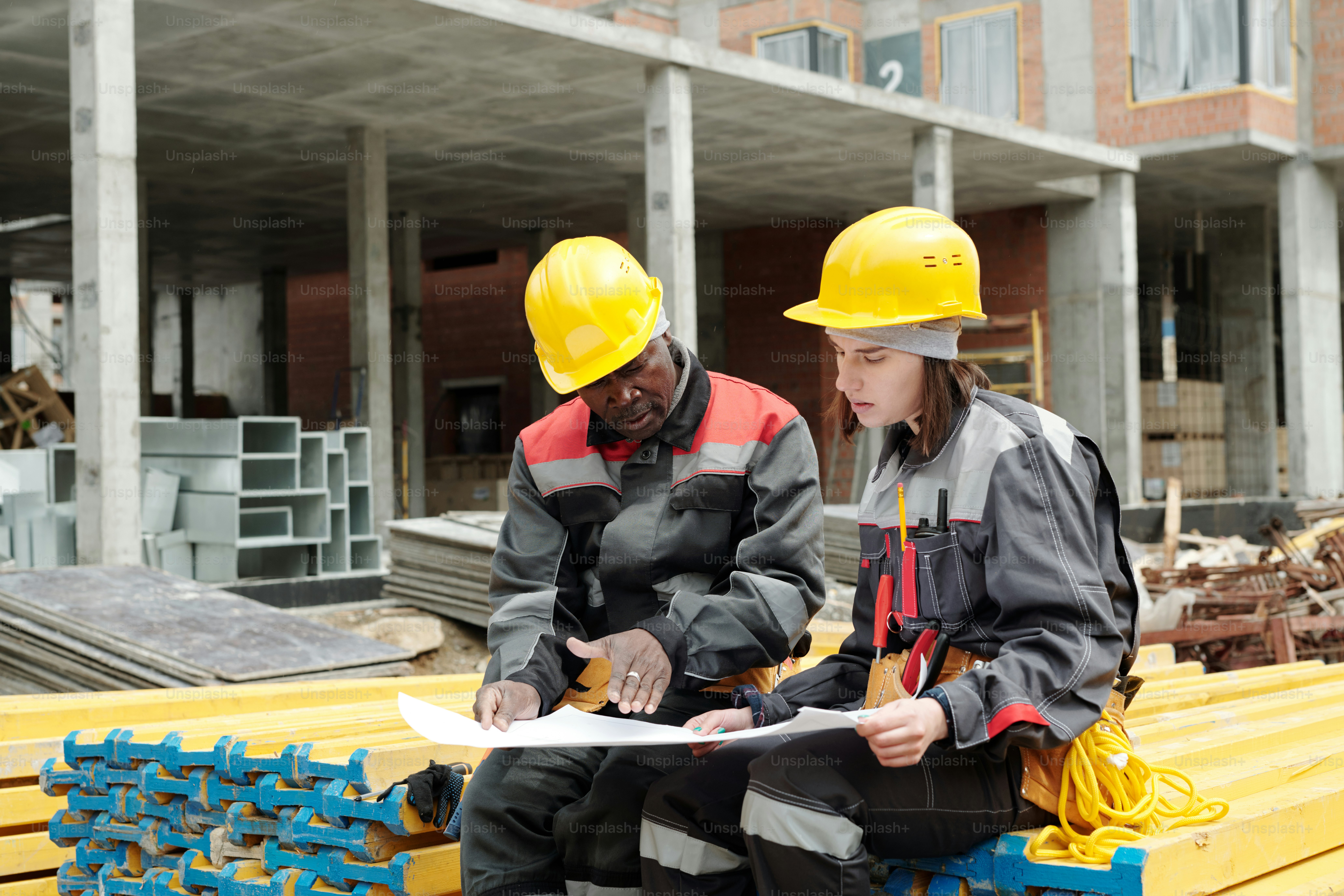 Young and mature builders in workwear and hardhats discussing sketch of ...