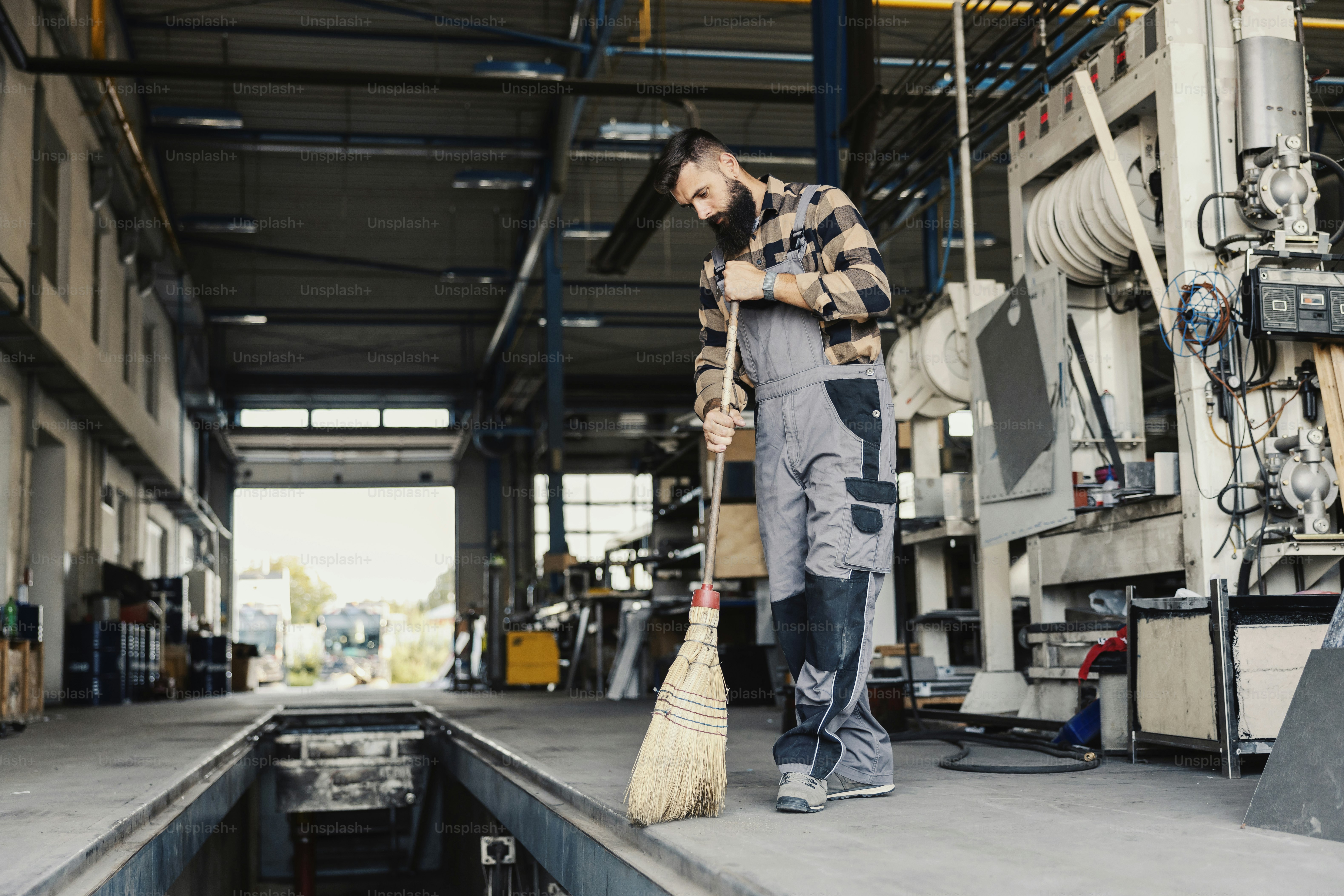A worker sweeping dust from the floor with broom in a mechanic's shop ...