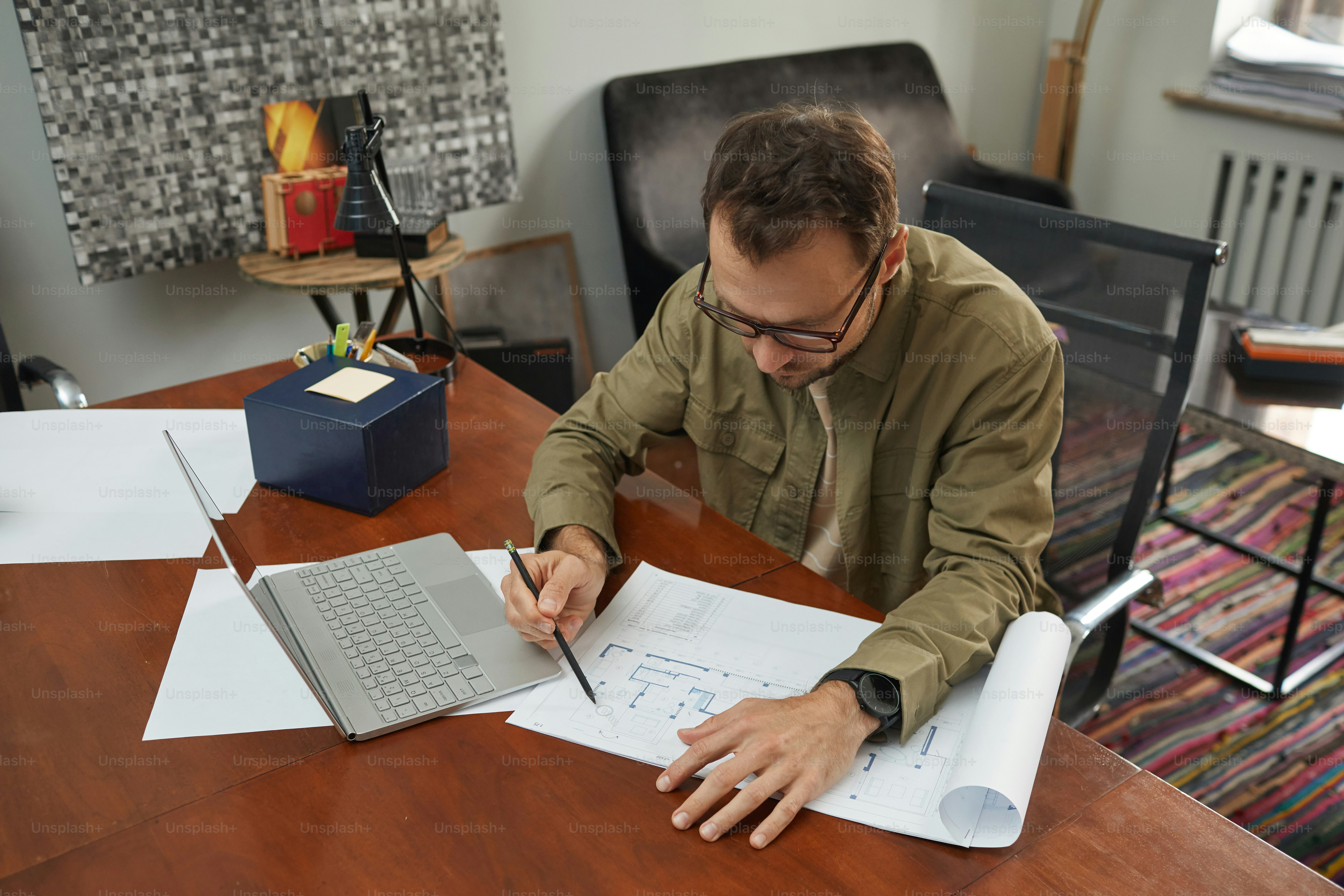Young architect in eyeglasses sitting at his workplace and examining ...