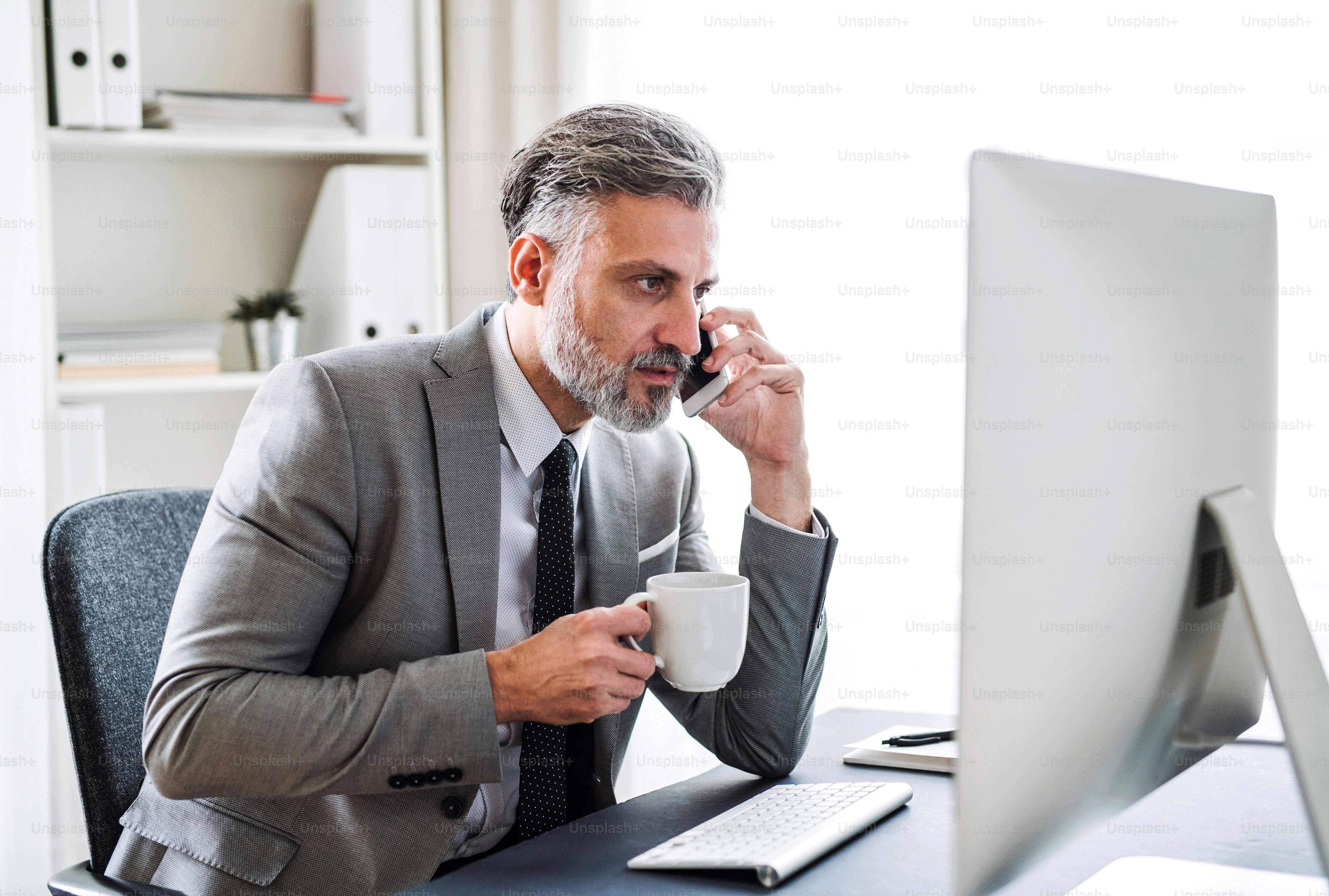 Serio uomo d'affari maturo con smartphone, caffè e computer desktop ...