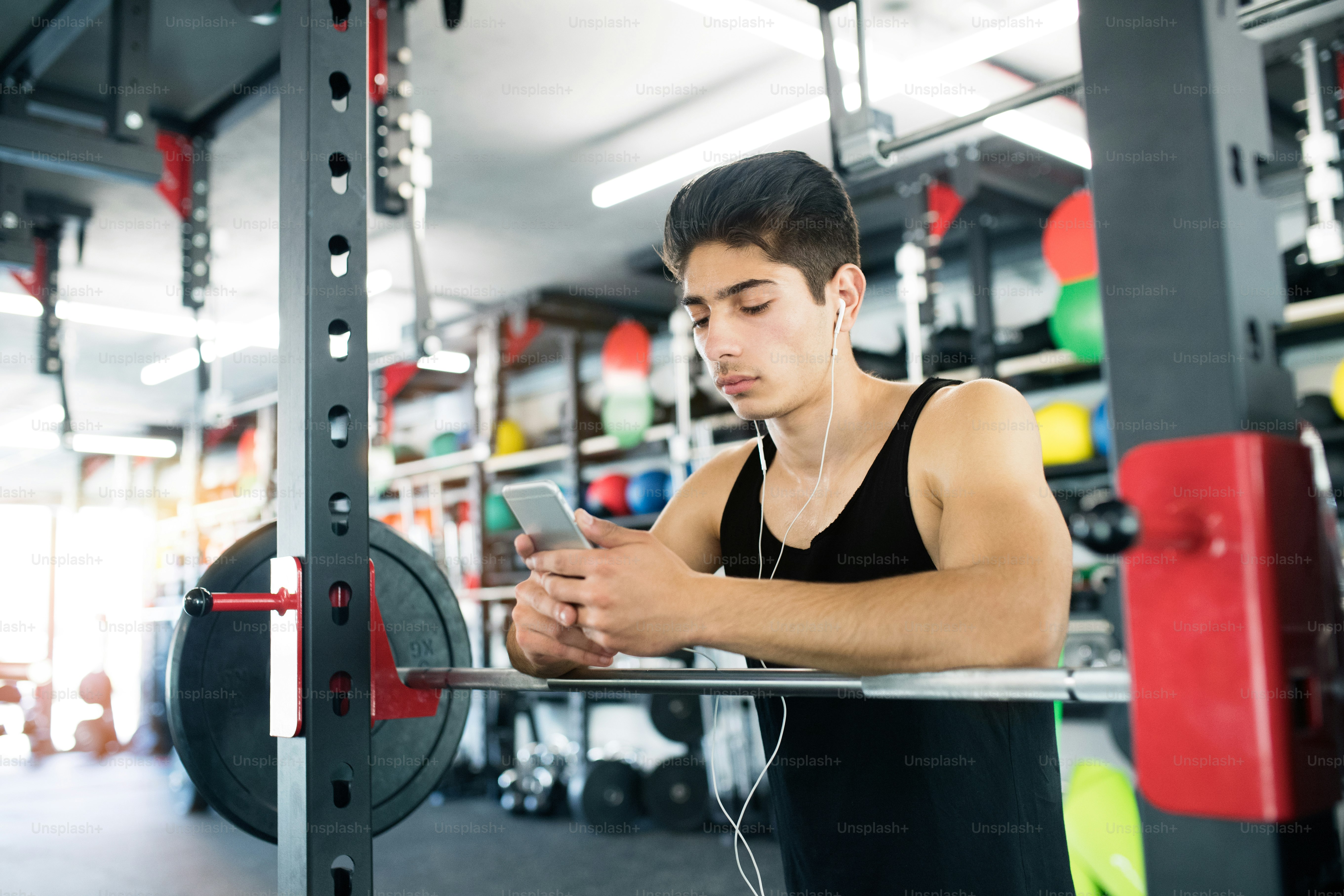 Hispanic fitness man in gym resting, holding smart phone, earphones in ...