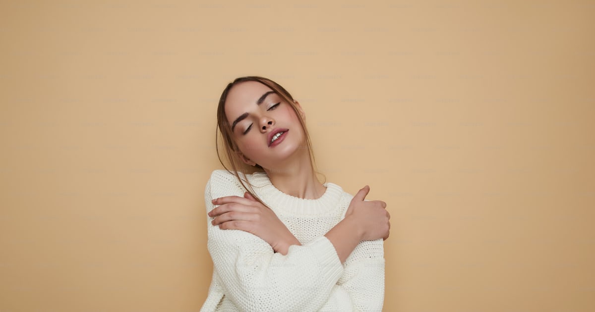 하얀 스웨터를 입은 어린 소녀가 베이지색 배경에 고립되어 서서 어깨에 손을 얹고 눈을 감고 있다 사진 Unsplash의