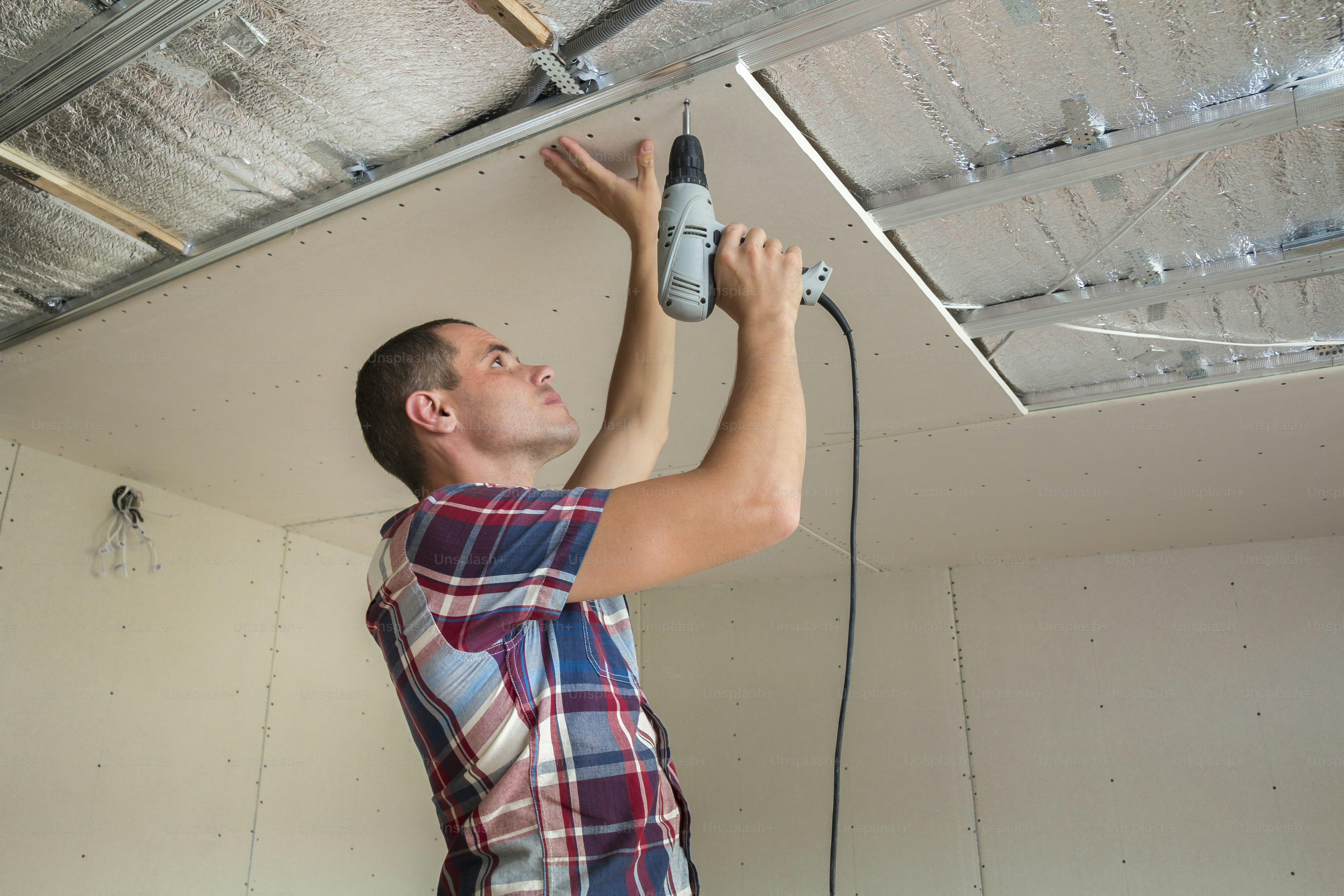 Young man in usual clothing and work gloves fixing drywall suspended ...