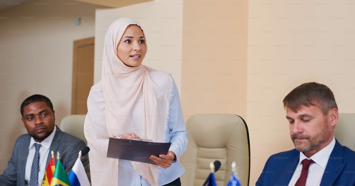 ヒジャーブを着た若いイスラム教徒の女性が、ホールの聴衆の前に立ちながら会議で報告をするの写真 ヒジャーブ Unsplashの写真