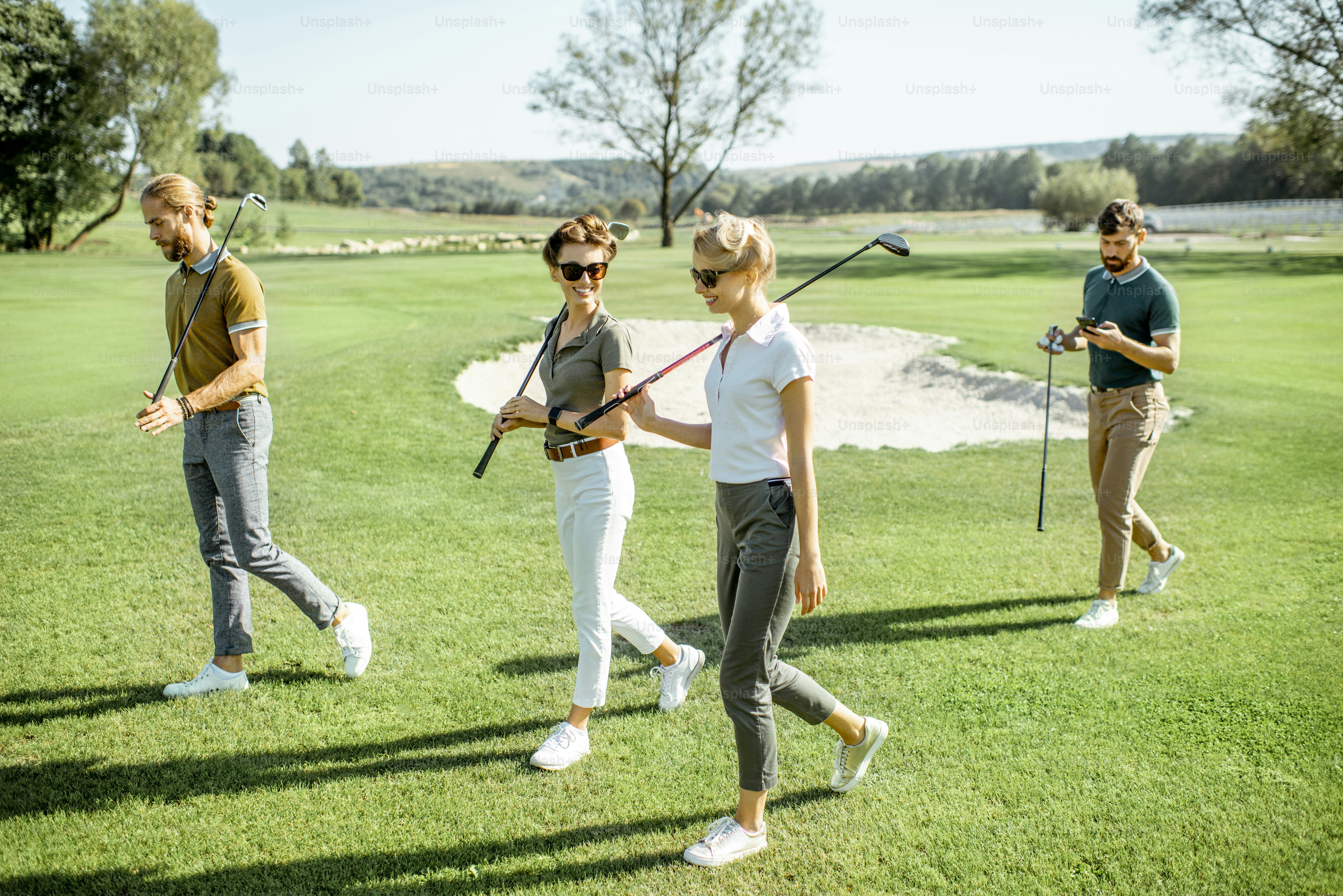 Young friends dressed casually walking with putters on the golf course during a game on a sunny ...