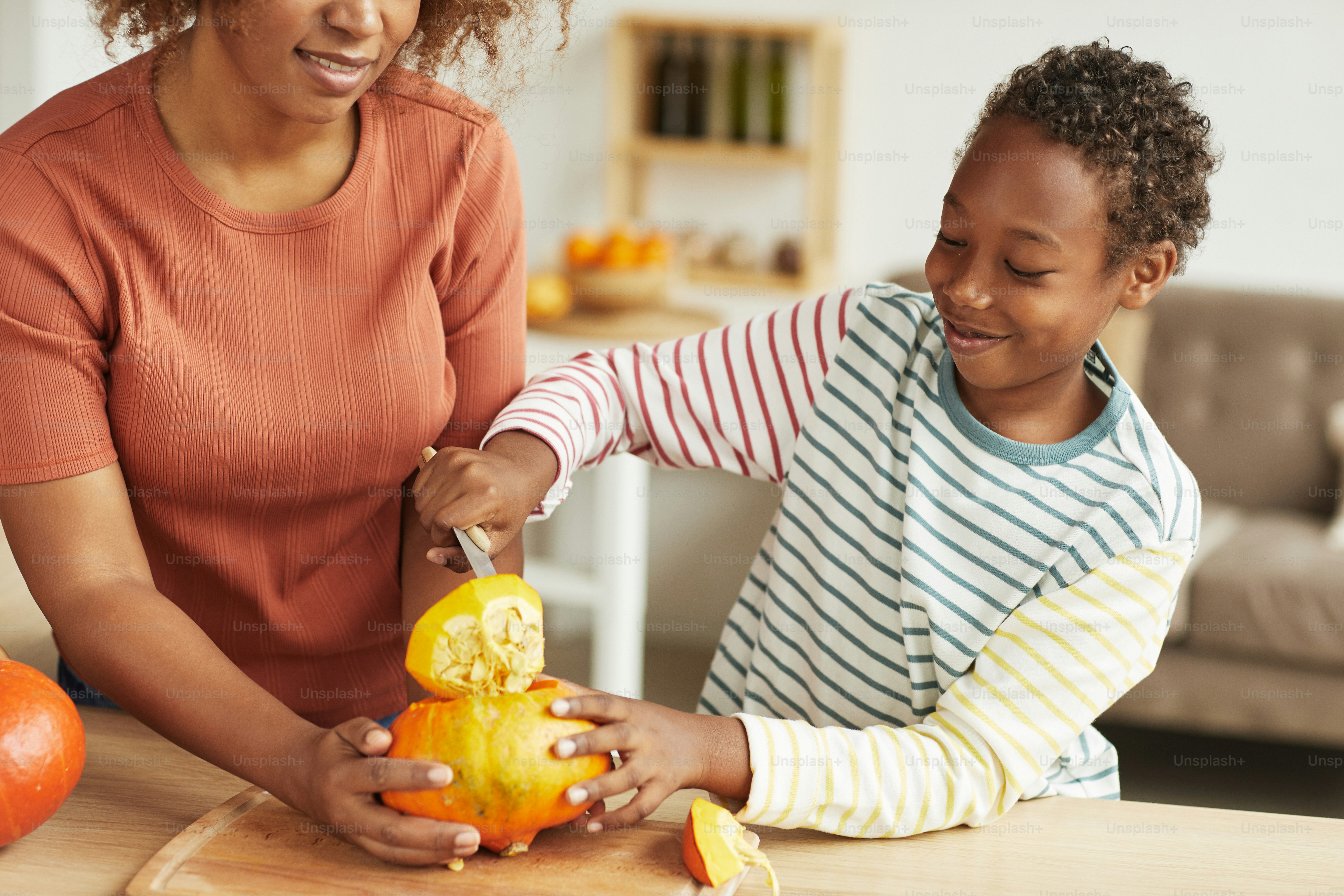 Cheerful African American boy cutting off pumpkin crown. his mother ...