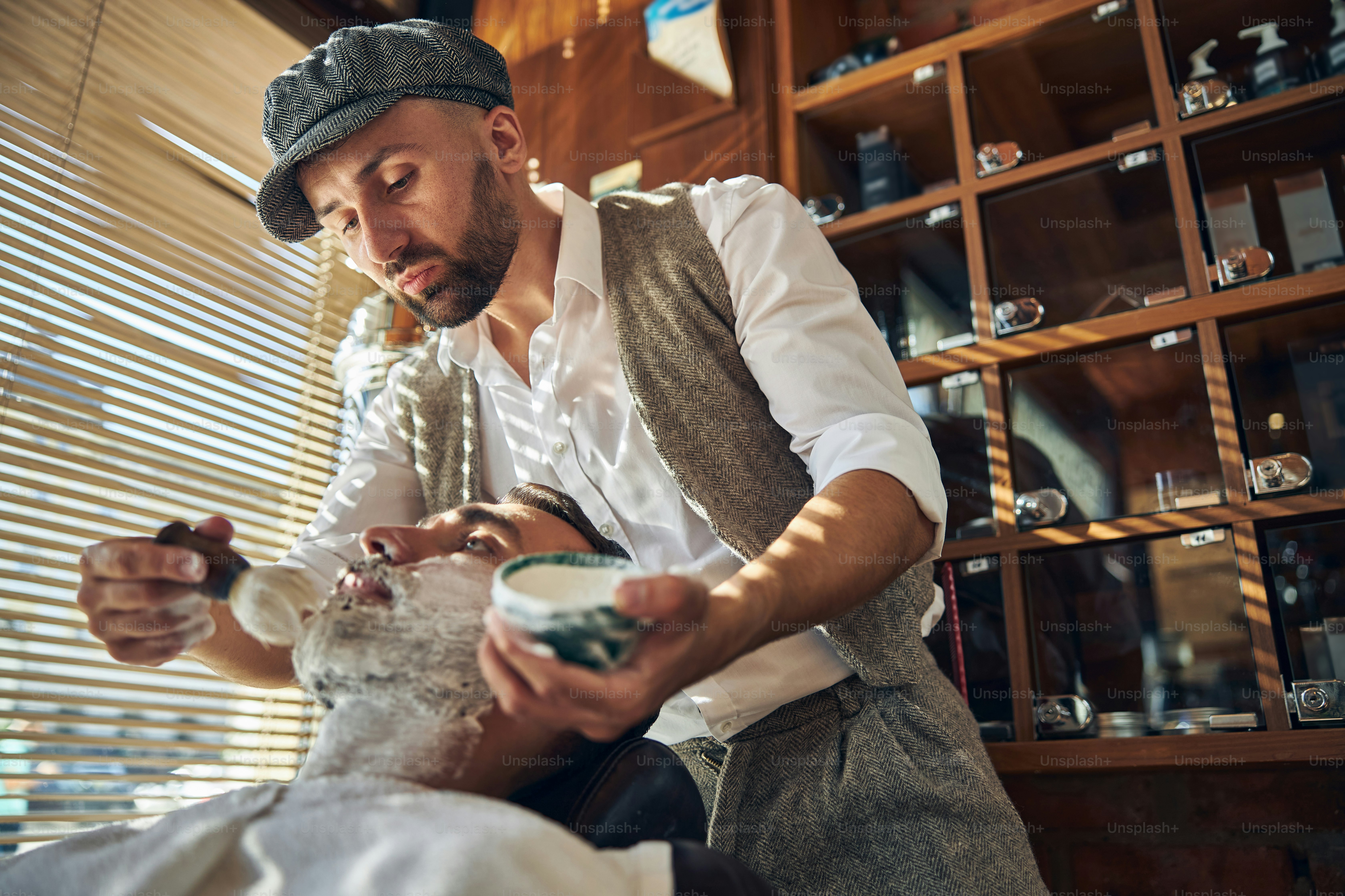 Focused barber in a peaky cap putting shaving cream all over the face ...