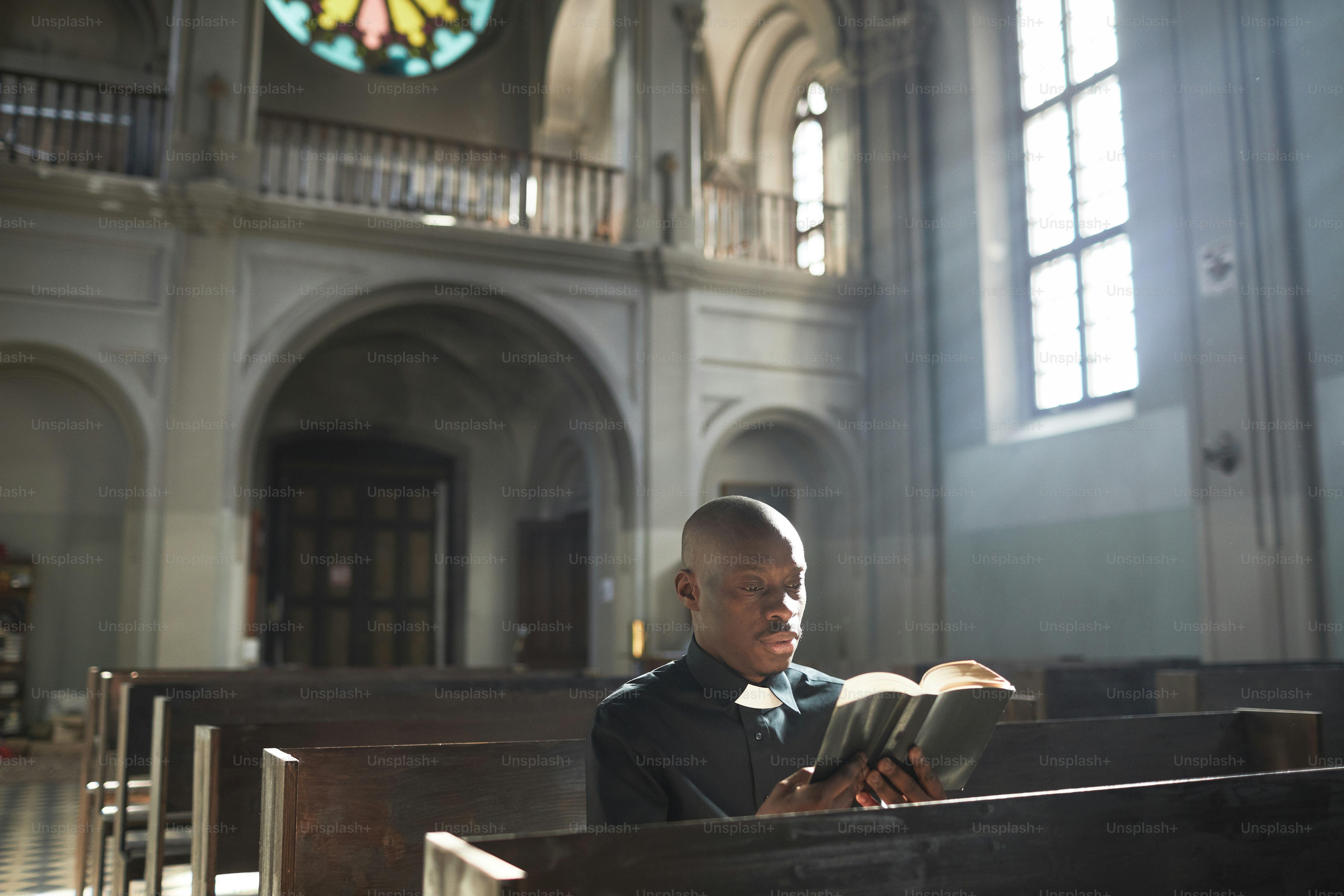 African priest in collar sitting on the bench in church and reading the ...