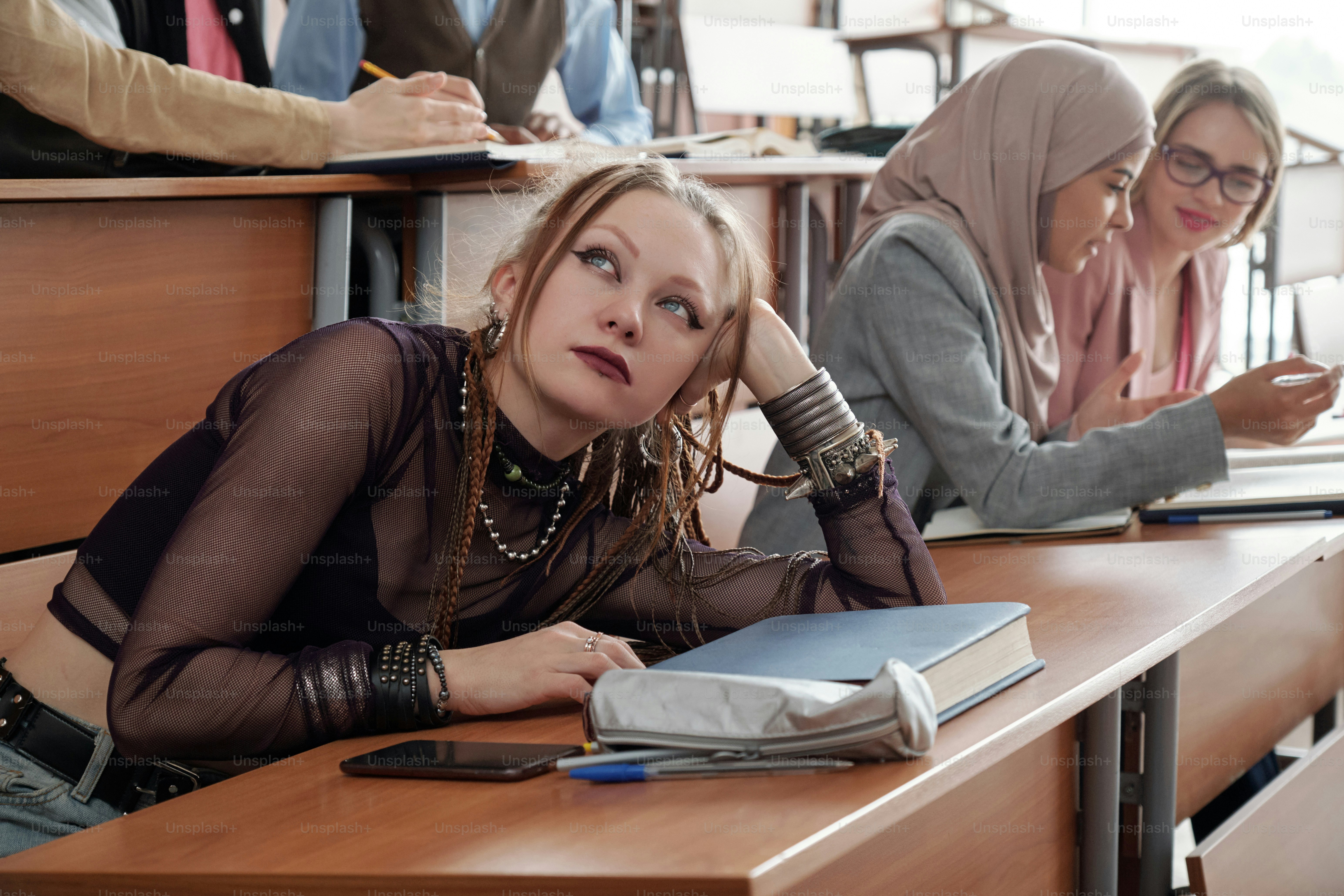 Distracted female teenager lying on desk at lesson while others working ...