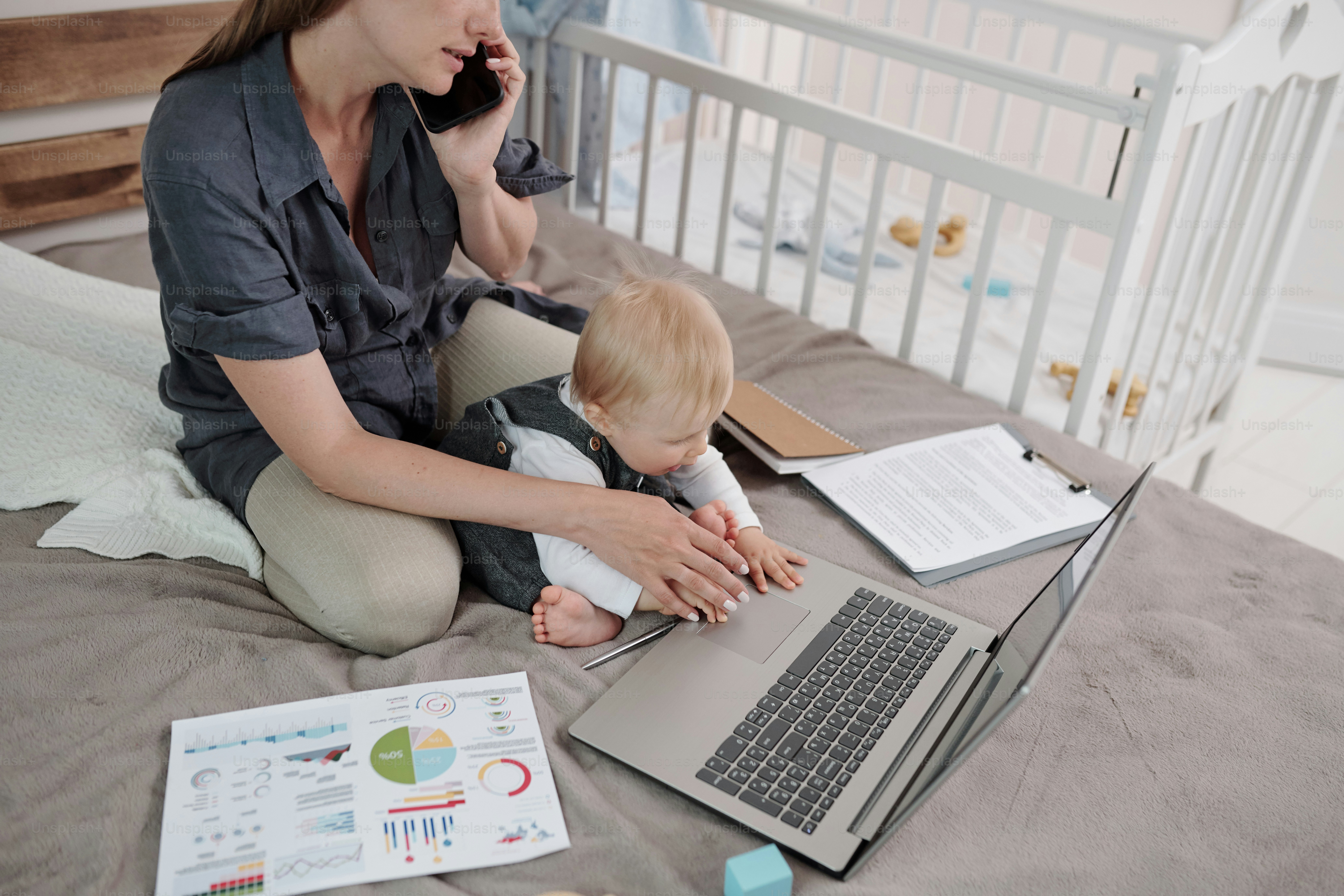 Multi-tasking young mother sitting with baby on bed and answering phone ...