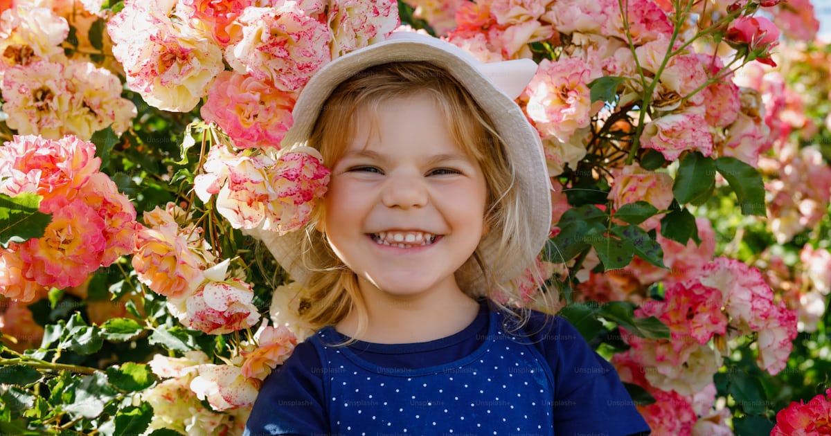 花の咲くバラ園の小さな幼児の女の子のポートレート。夏の晴れた日に公園でバラや花を楽しむかわいい美しい素敵な子。幸せな笑顔の赤ちゃんの写真