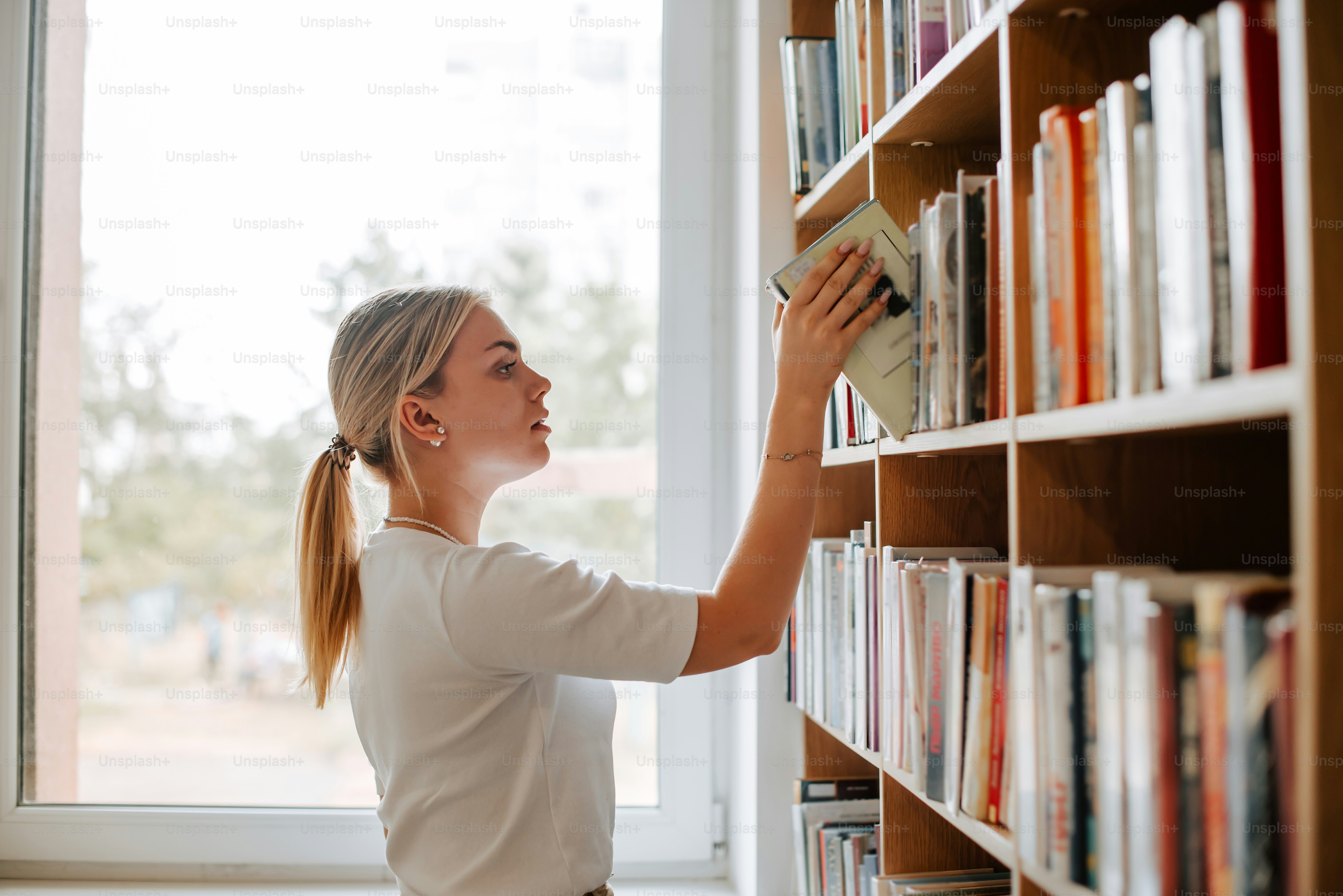 A woman is picking up a book from a book shelf photo – College Image on ...