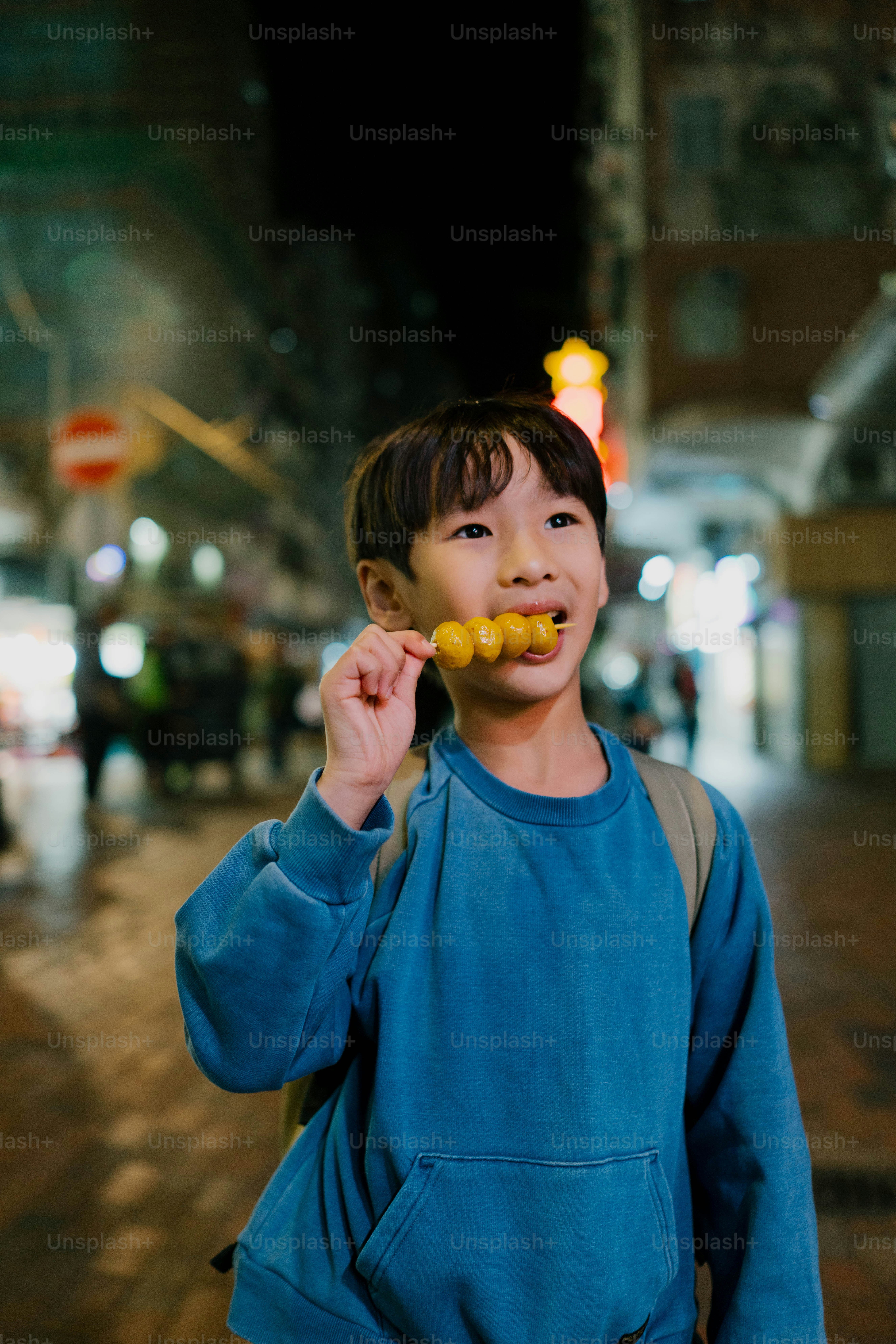 A young boy standing on a sidewalk holding a piece of food in his mouth ...