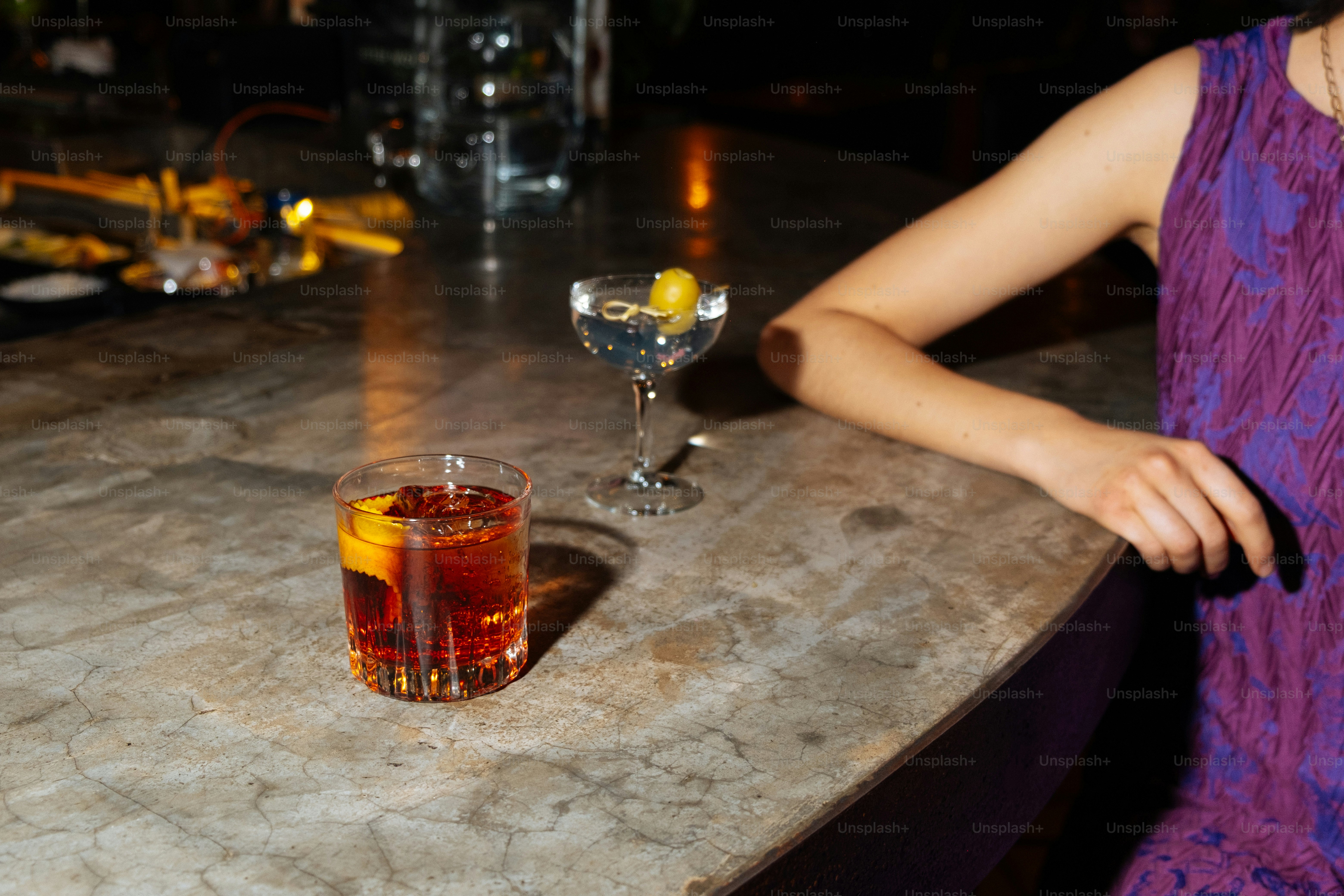 Cocktails rest on a bar alongside a person. photo – Bar Image on Unsplash